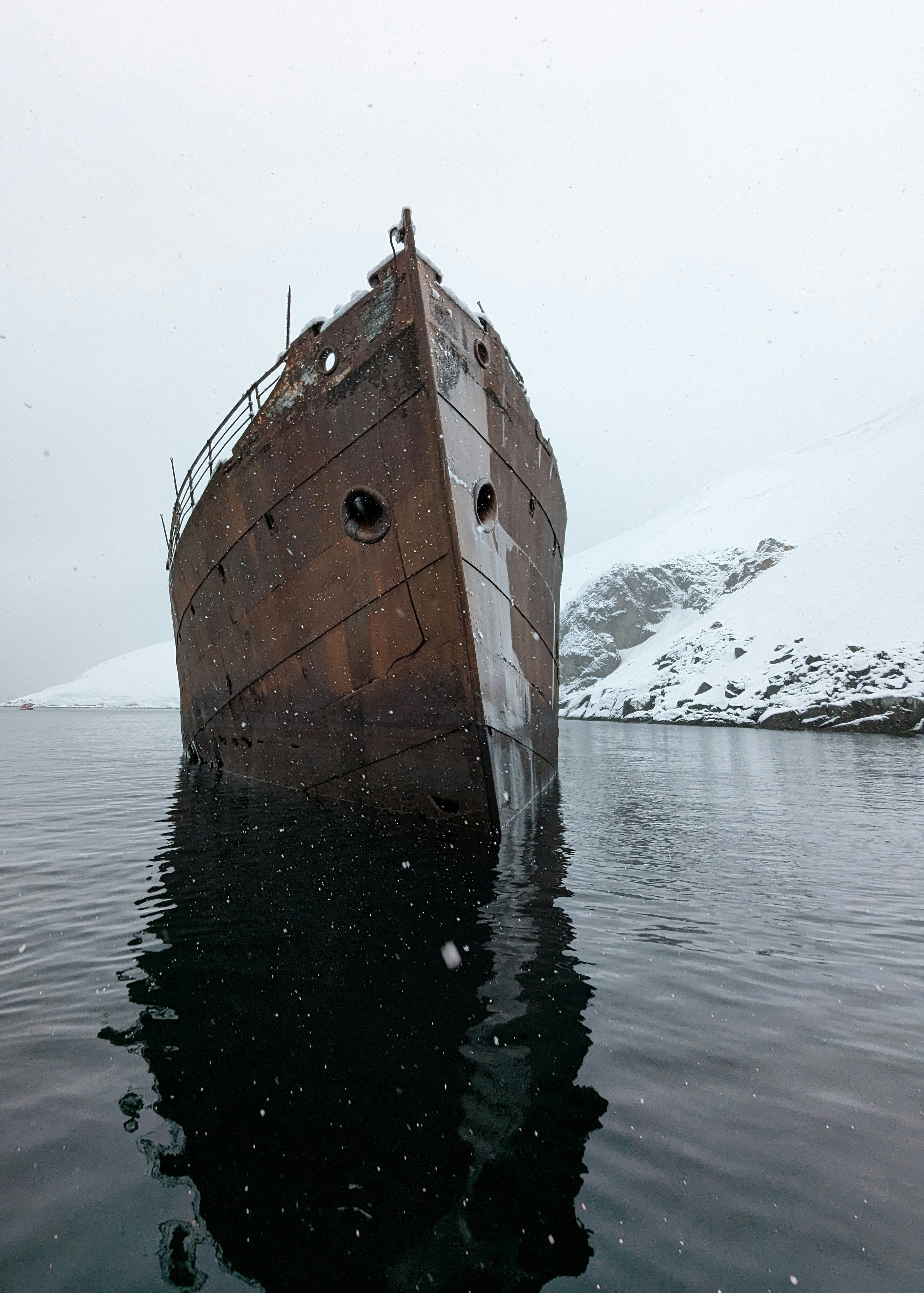 Photograph of a rusted shipwreck rising from calm, dark water with snow-covered cliffs in the background.