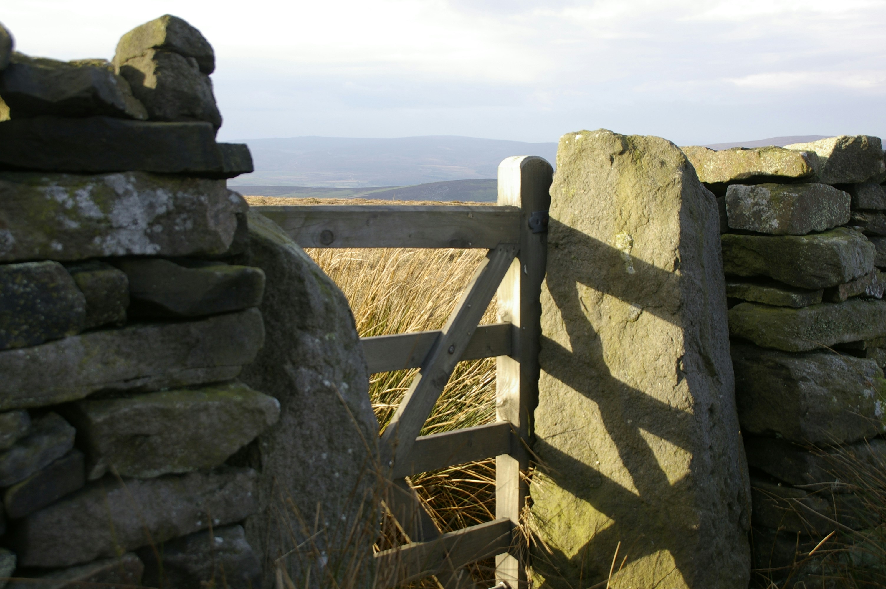 Weathered stone gate and wall frame a distant hillside and grasses, suggesting a highland path opening to a wide view.