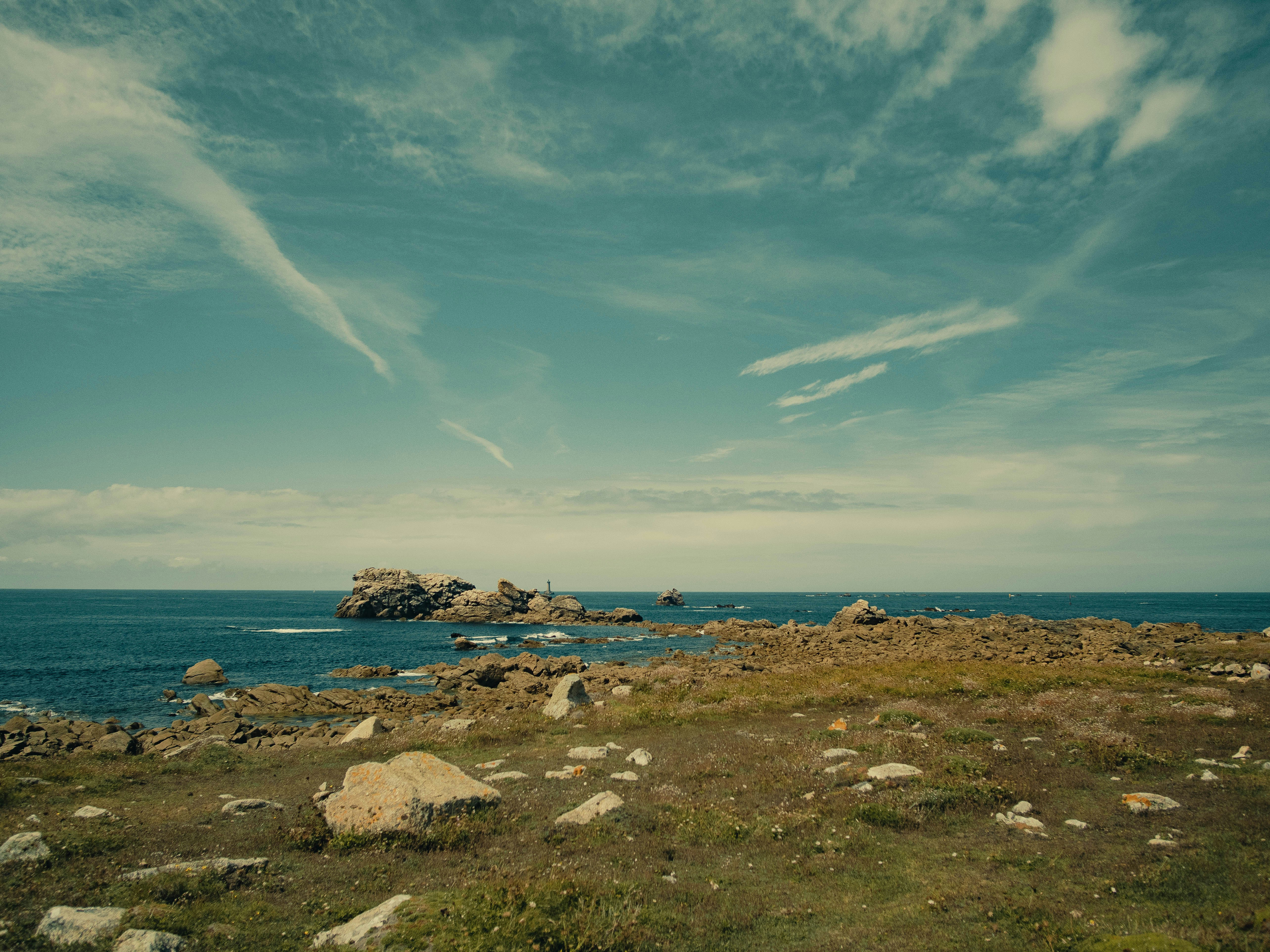 Rocky coastline under a vast sky with wispy clouds, featuring a distant rock formation emerging from the sea.