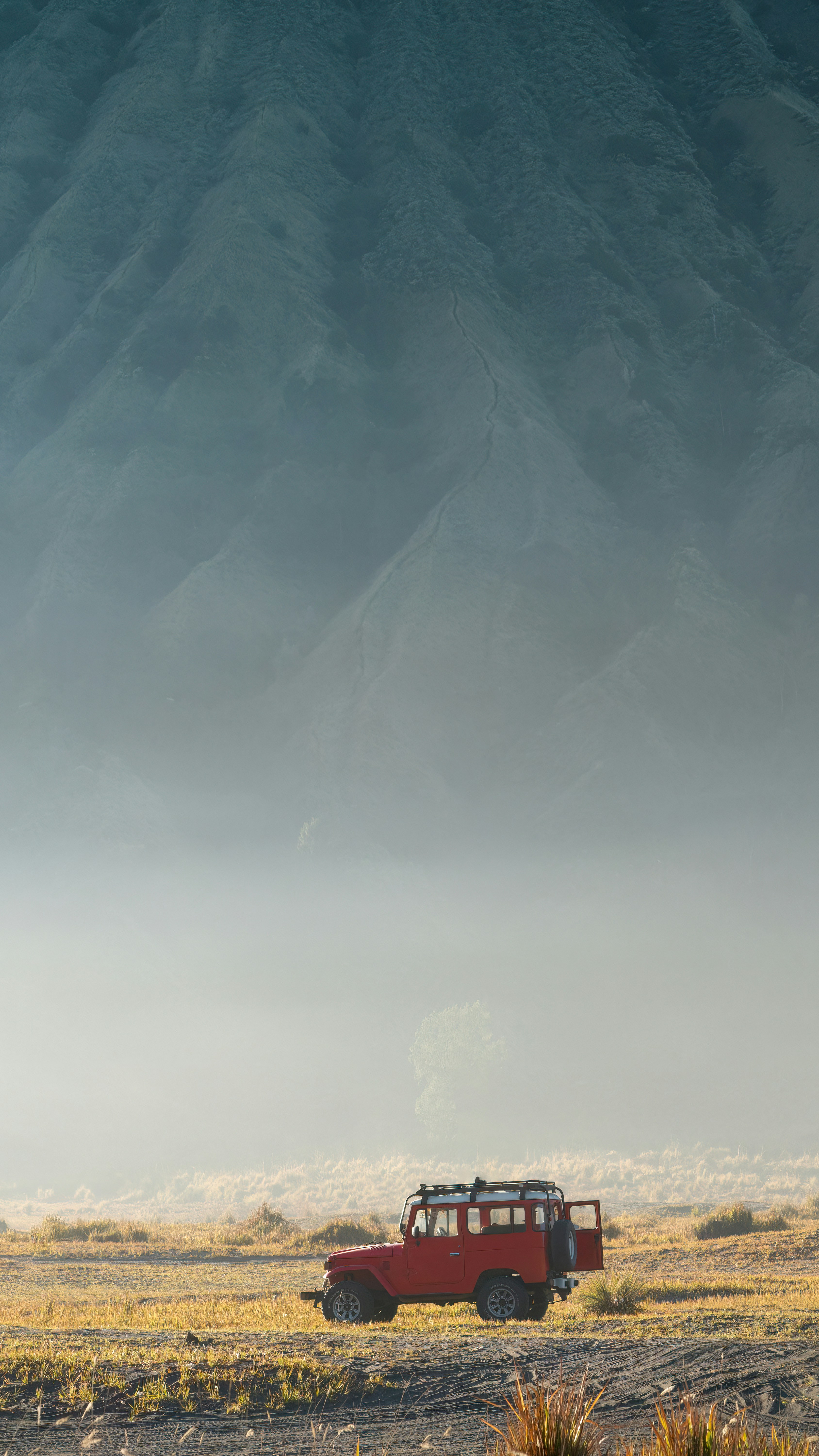 A vintage red jeep parked in a foggy landscape, surrounded by rolling hills and a solitary tree in the background.