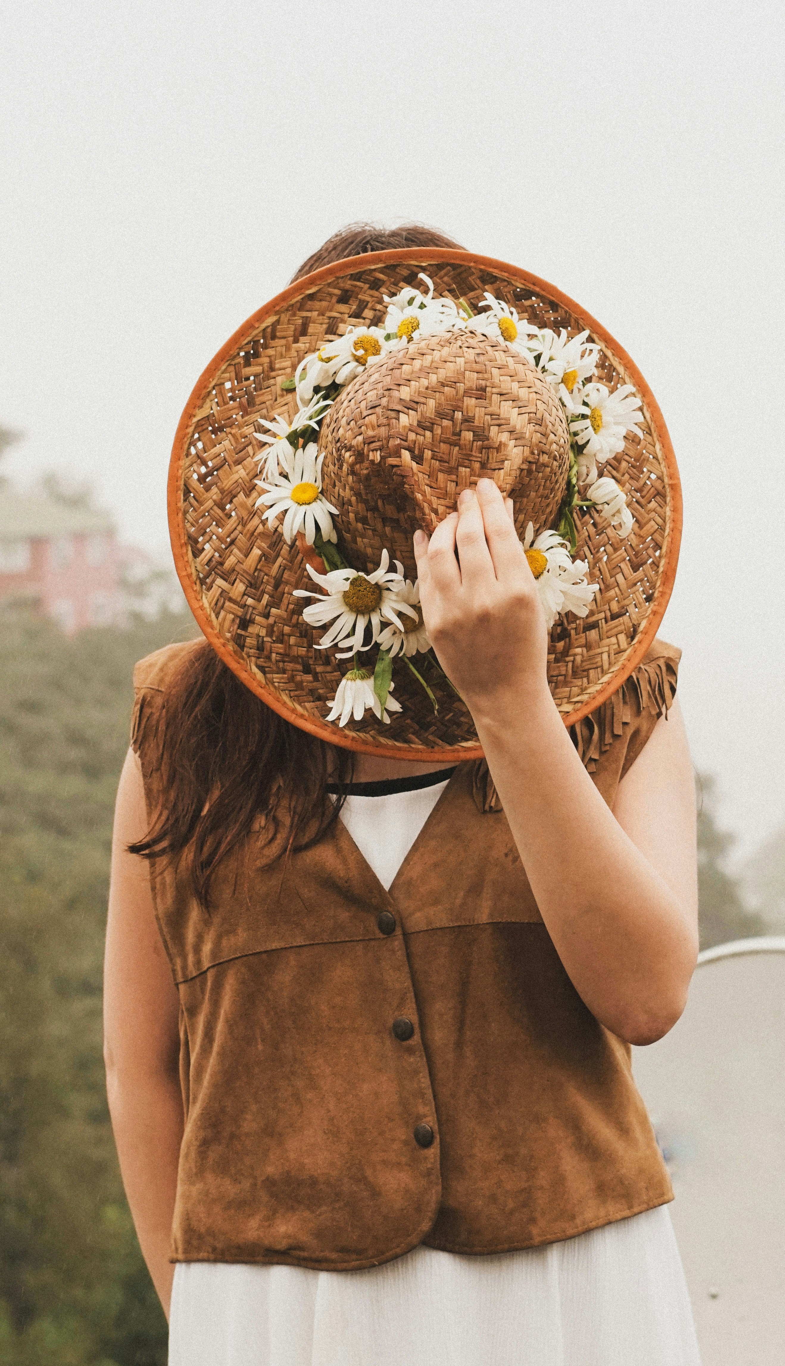 Photograph of a person outdoors wearing a wide straw hat decorated with daisies, face hidden by the hat, with a warm, vintage mood.