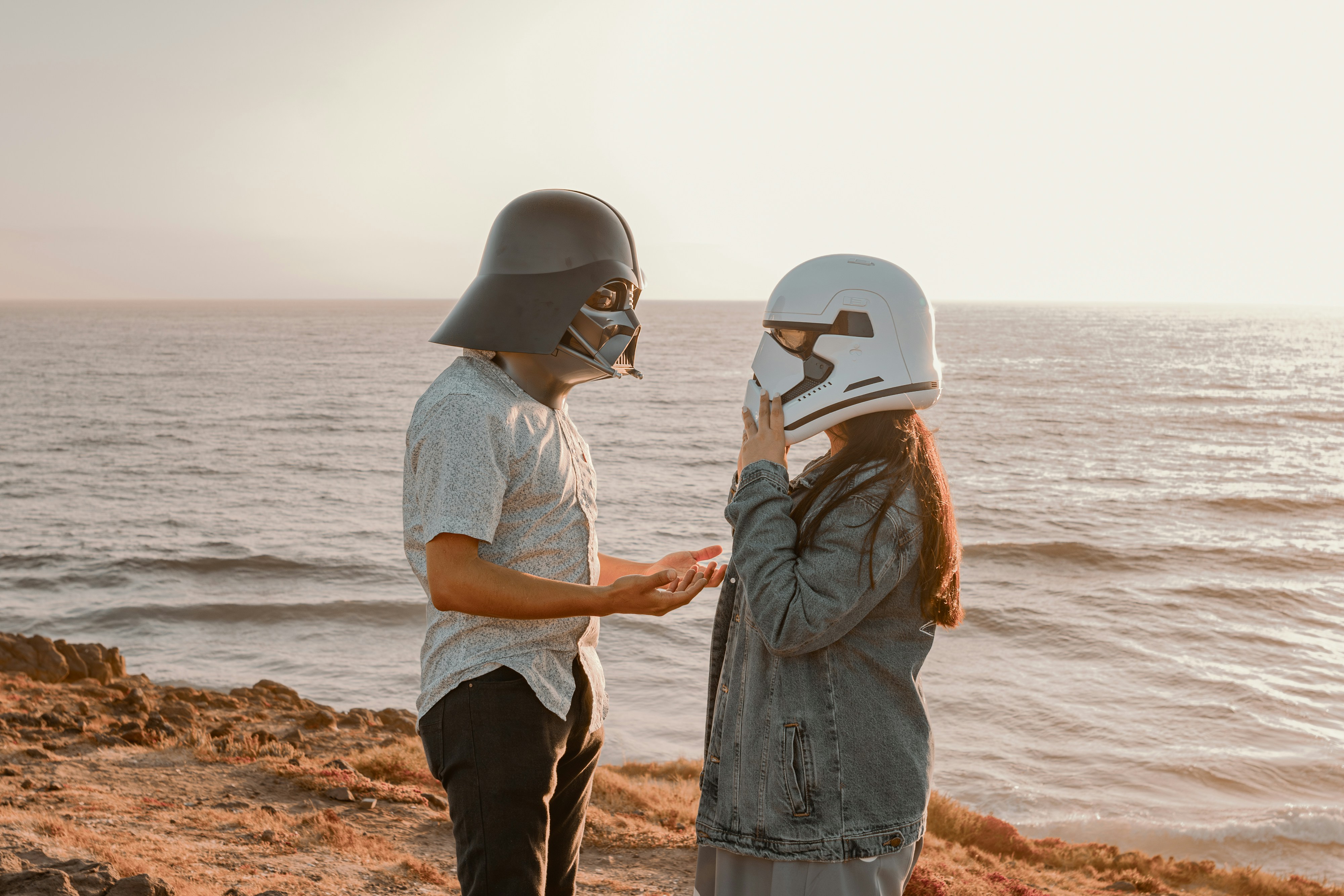 Two individuals wearing helmets converse on a rocky shoreline as the sun sets over the calm sea.