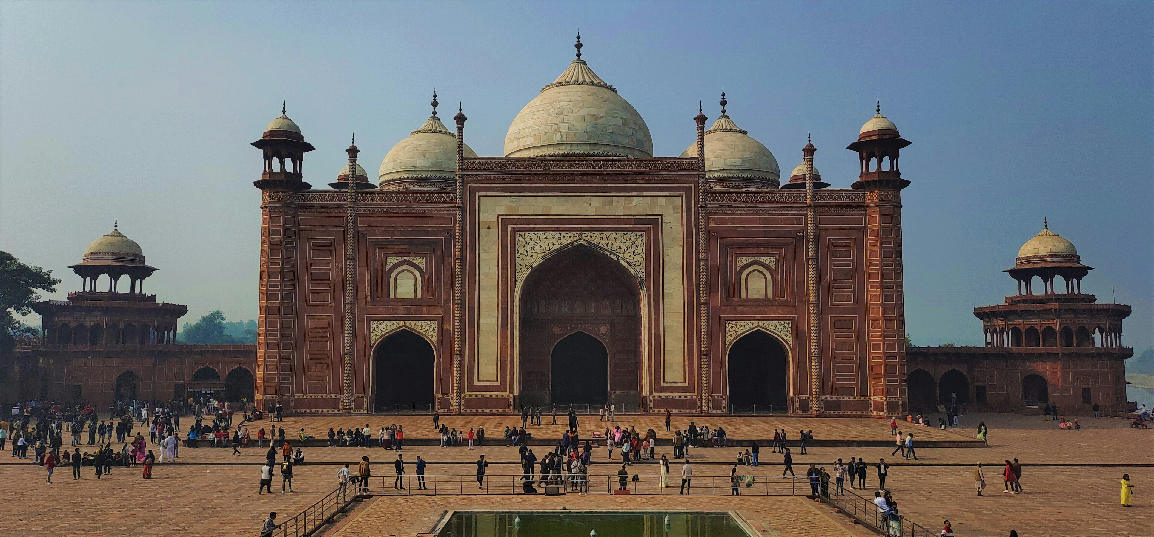 Intricate red sandstone architecture of a historical monument, featuring majestic domes and detailed carvings, bustling with visitors. A serene reflecting pool enhances the scene.