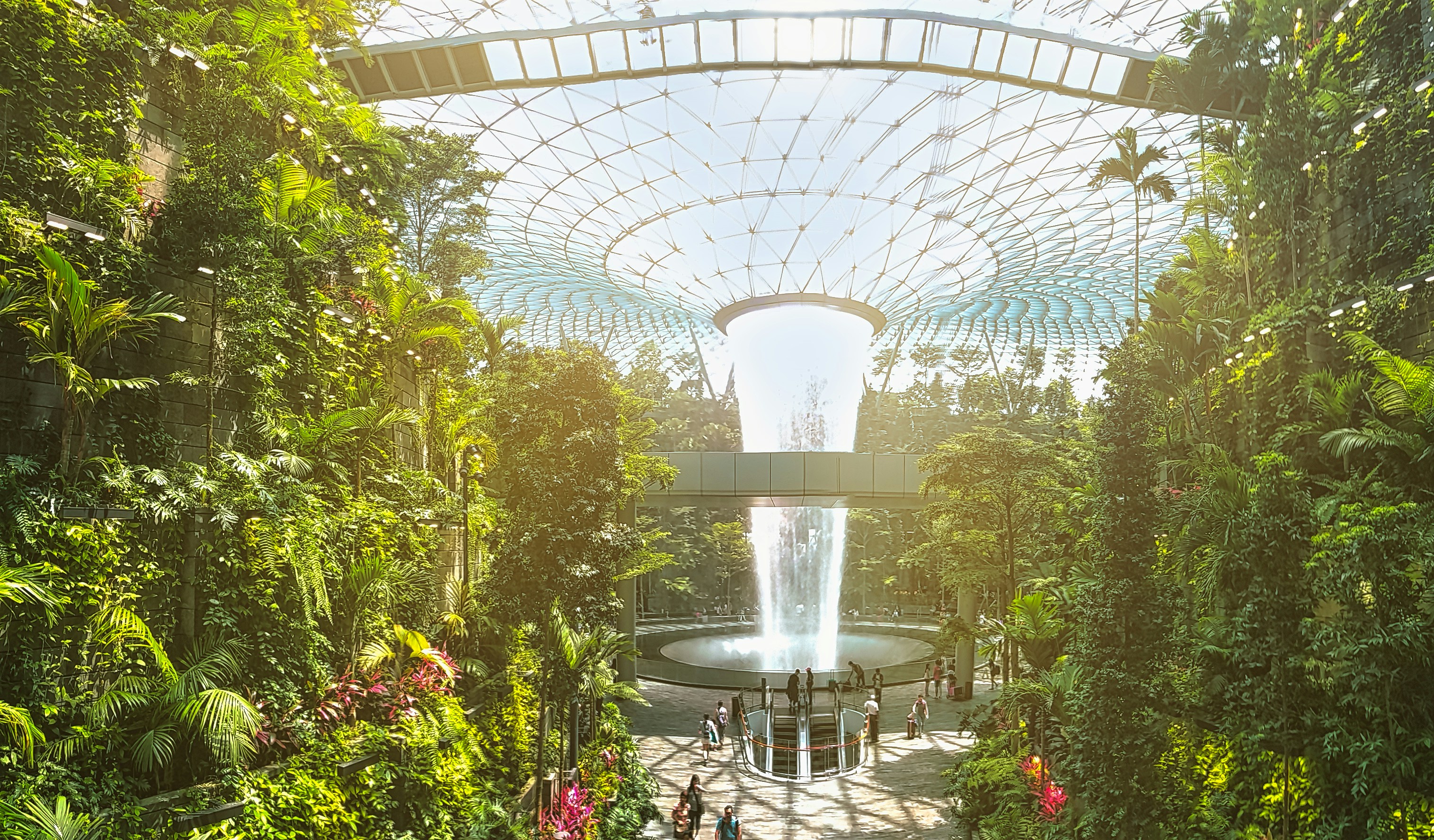 Inside a glass-domed conservatory, a tall central fountain rises above a circular pool. Lush tropical plants line the atrium as visitors stroll the sunlit walkway.