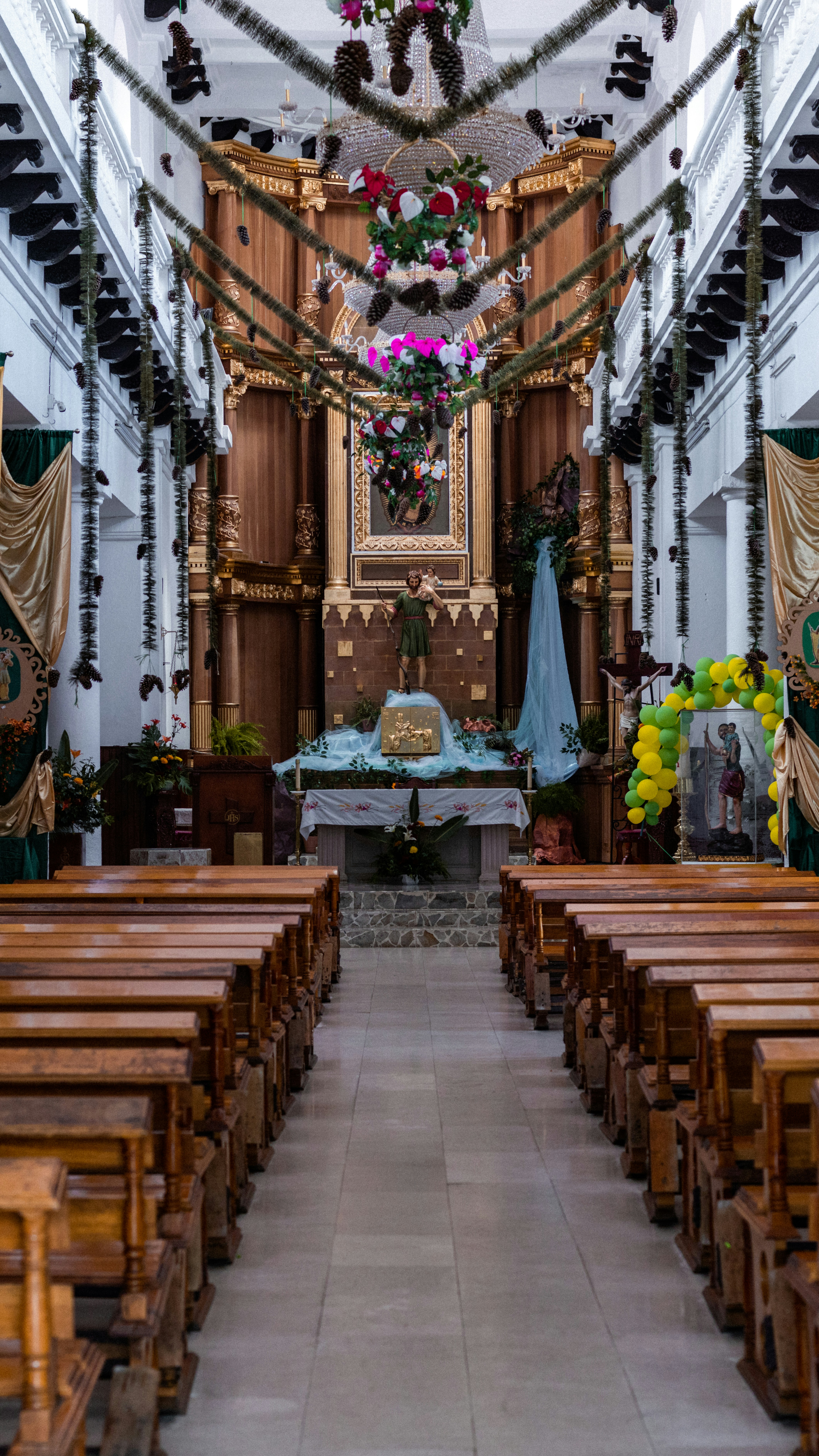 Interior of a church with rows of wooden pews guiding the eye toward a gilded altar adorned with flowers and drapery.