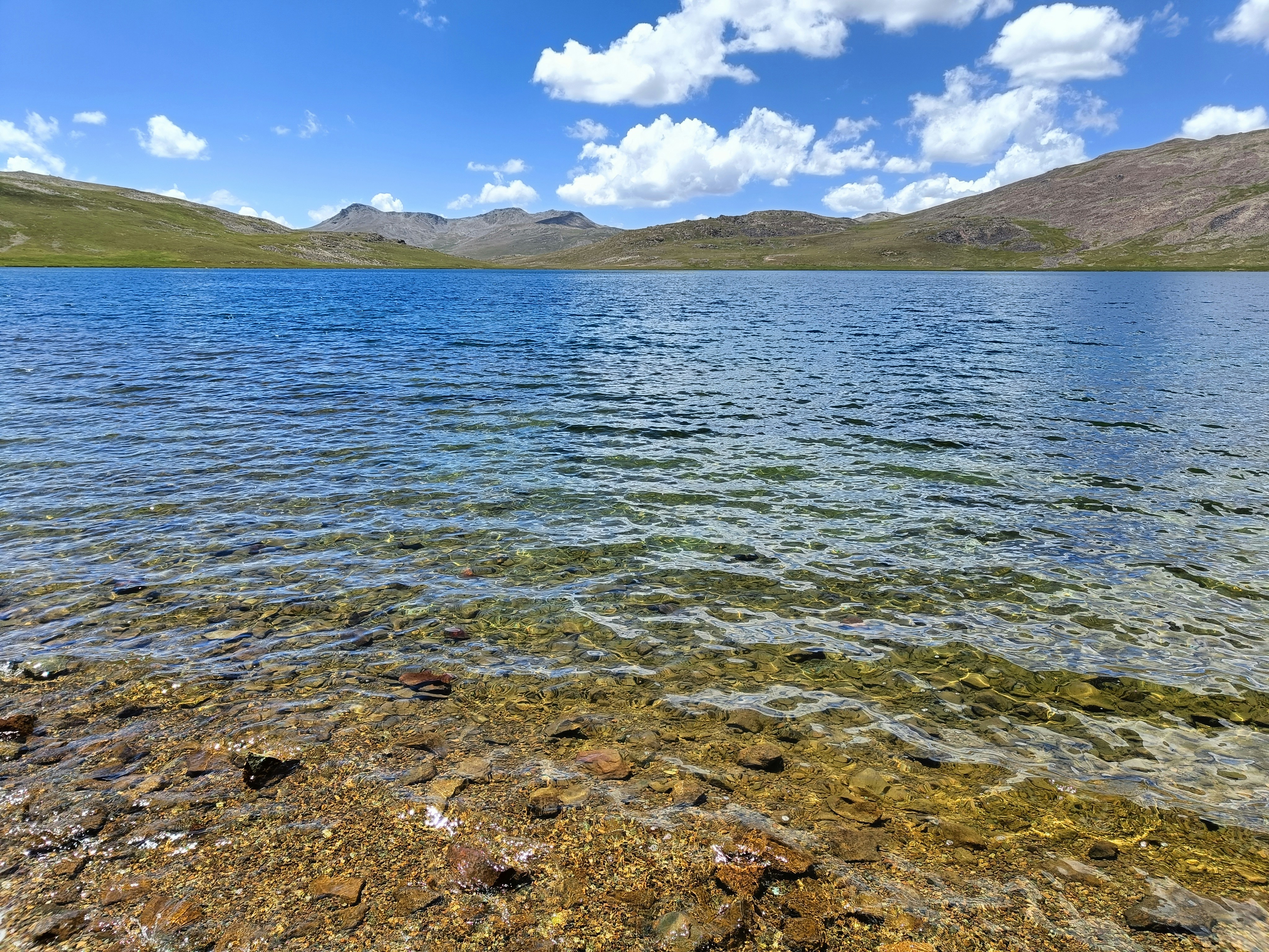 Crystal-clear mountain lake with a rocky shore and surrounding hills under a bright blue sky dotted with clouds.