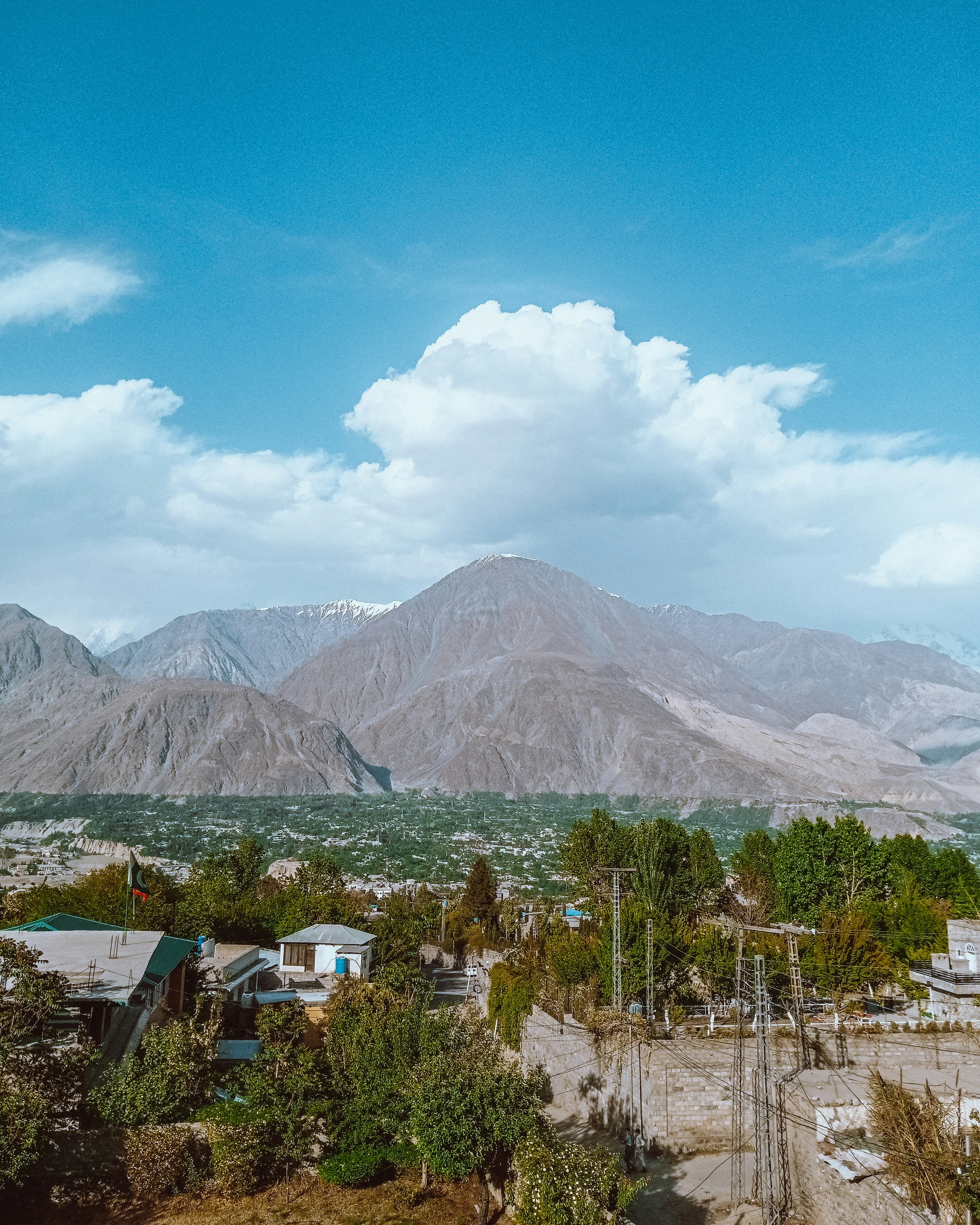 Landscape photograph of a mountain range looming behind a small village, under a bright blue sky with scattered clouds. The scene emphasizes the contrast between rugged highlands and the tranquil settlement below.