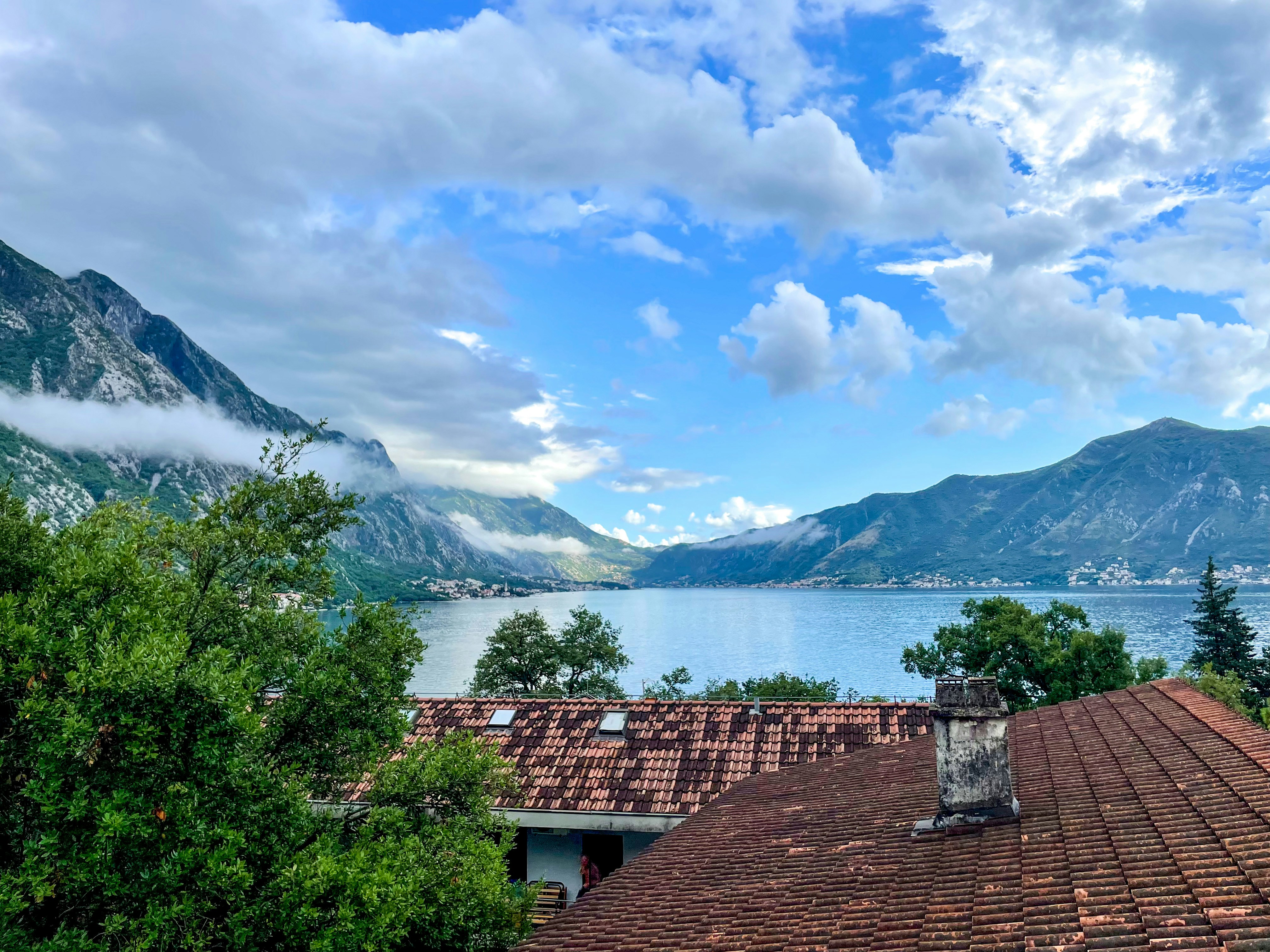 Mountains and a lake viewed beyond a red-tiled roof under a sky filled with fluffy clouds.