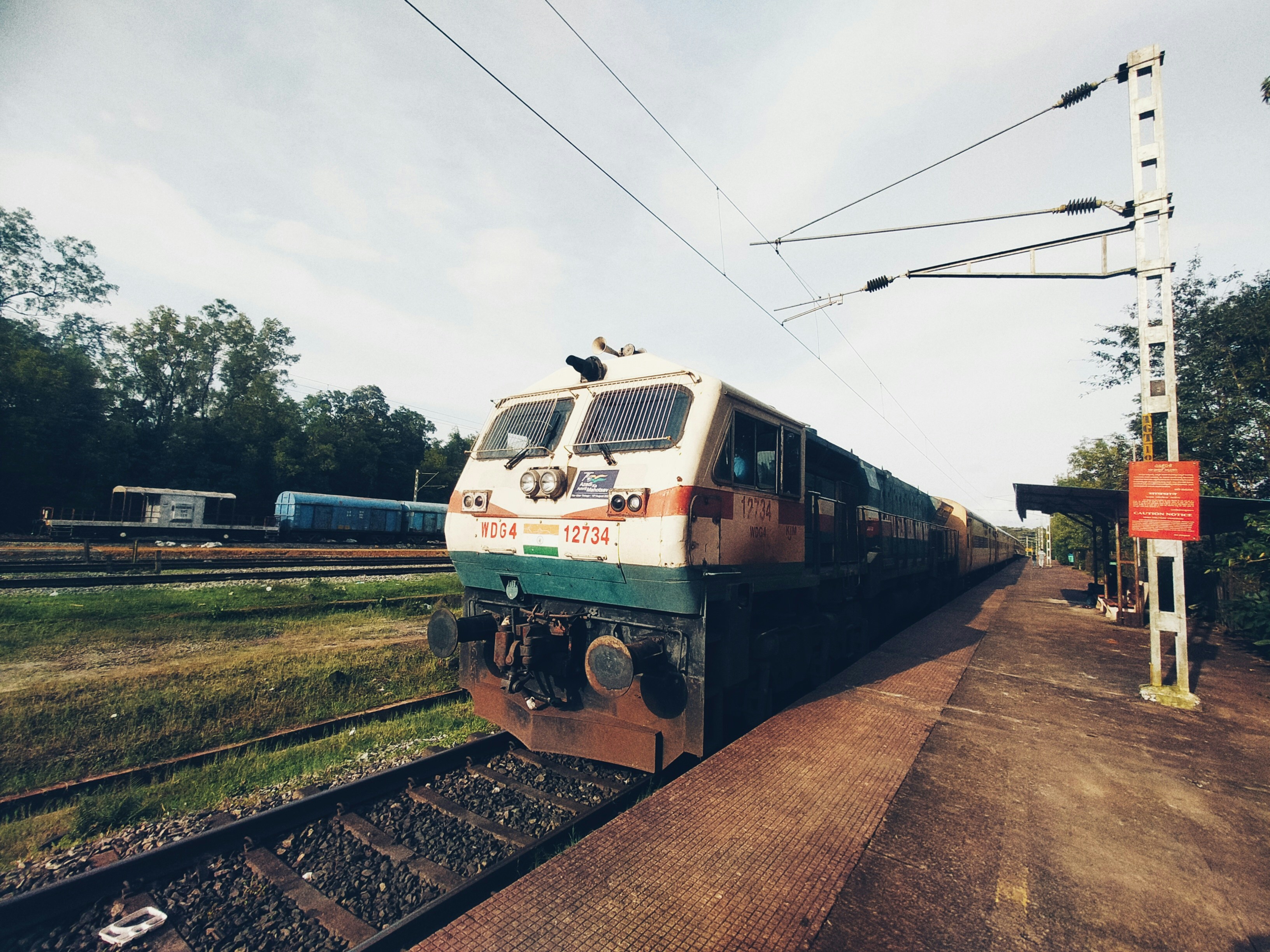 Train approaching a rural station surrounded by lush greenery under a bright sky.