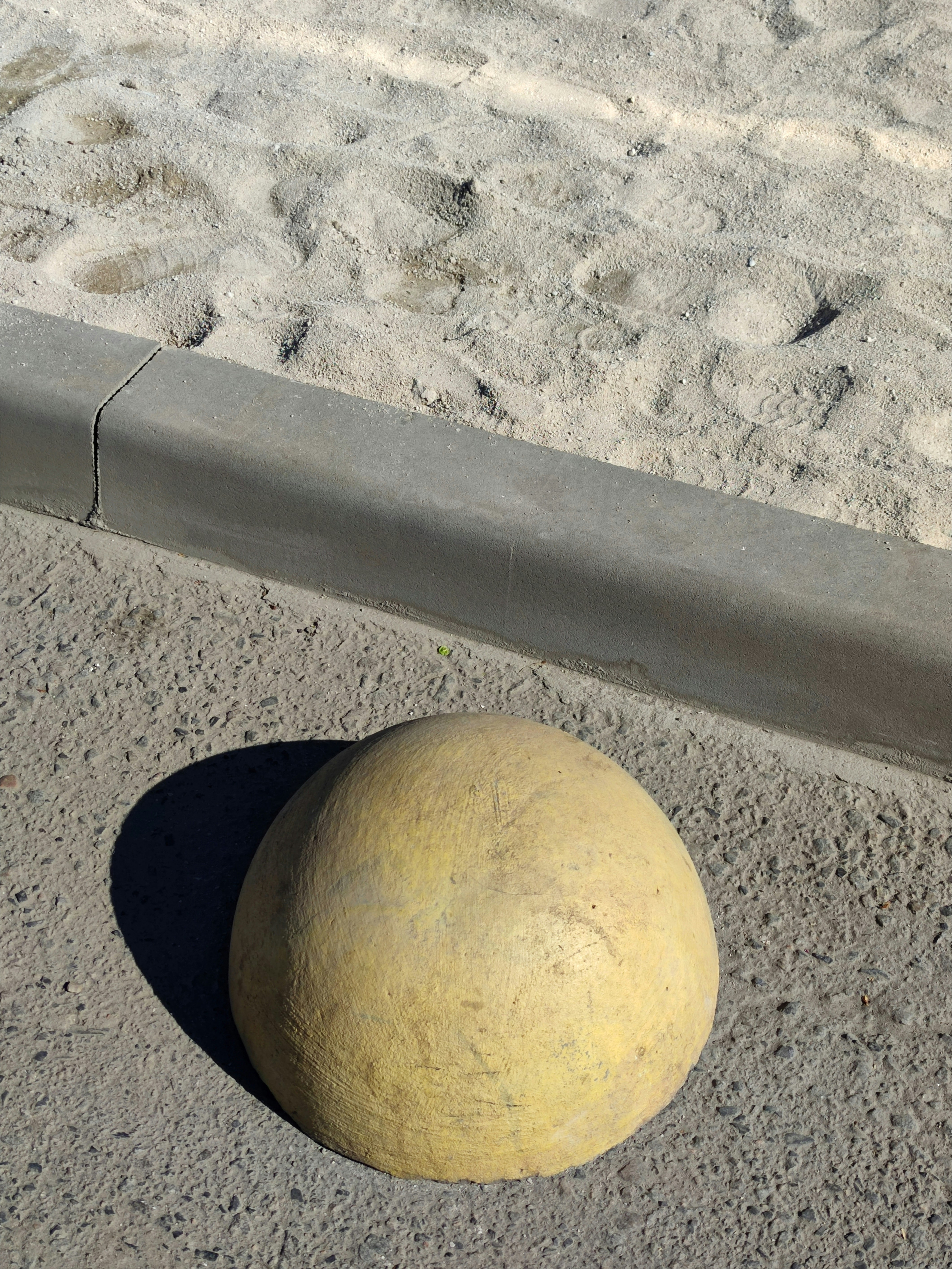 Yellow ball sits beside a concrete curb with sandy ground beyond, forming simple geometry in a quiet street-scene.
