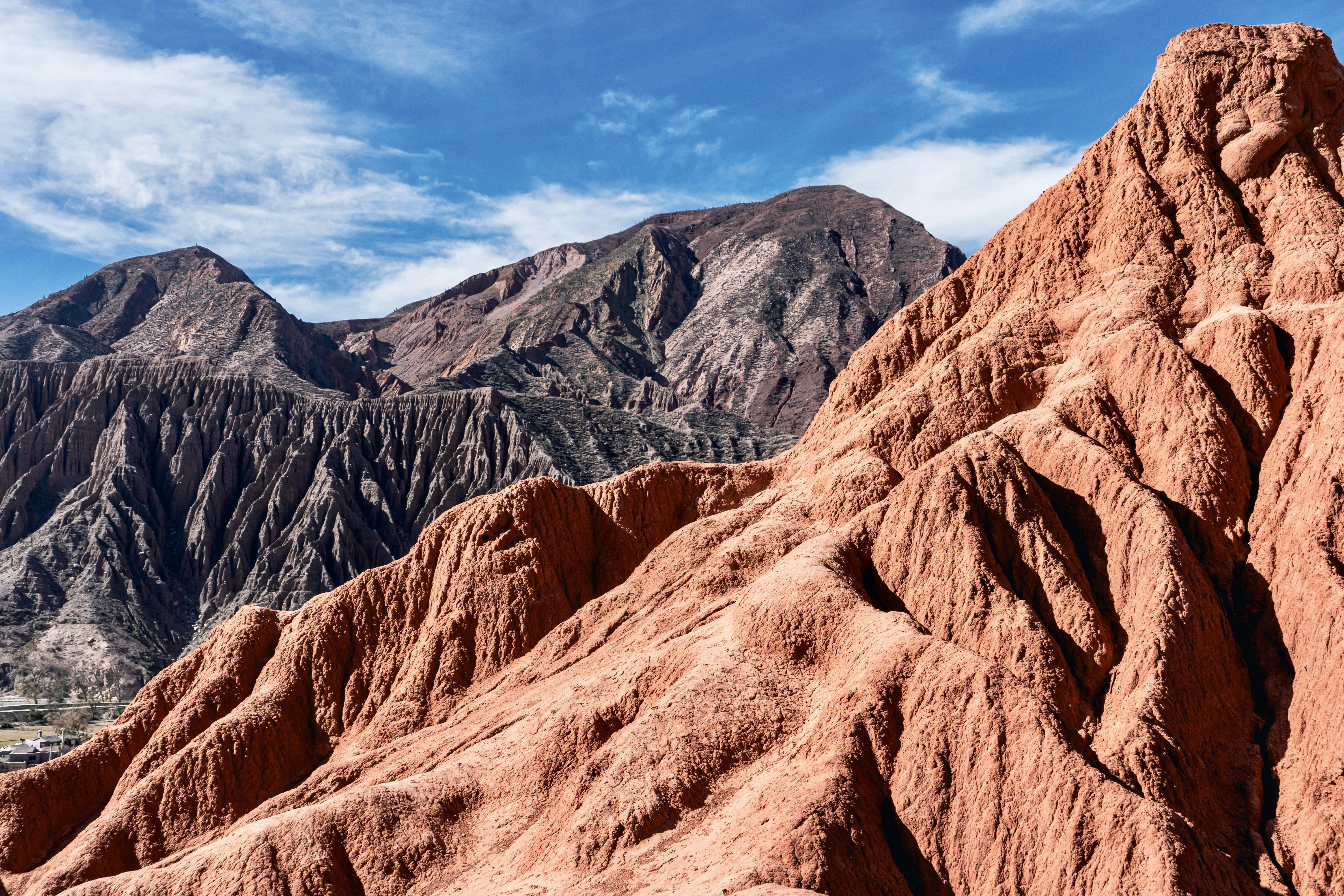 Dramatic red rock formations rise against a backdrop of rugged mountains under a bright blue sky. The textures and shadows reveal the intricate patterns shaped by nature.