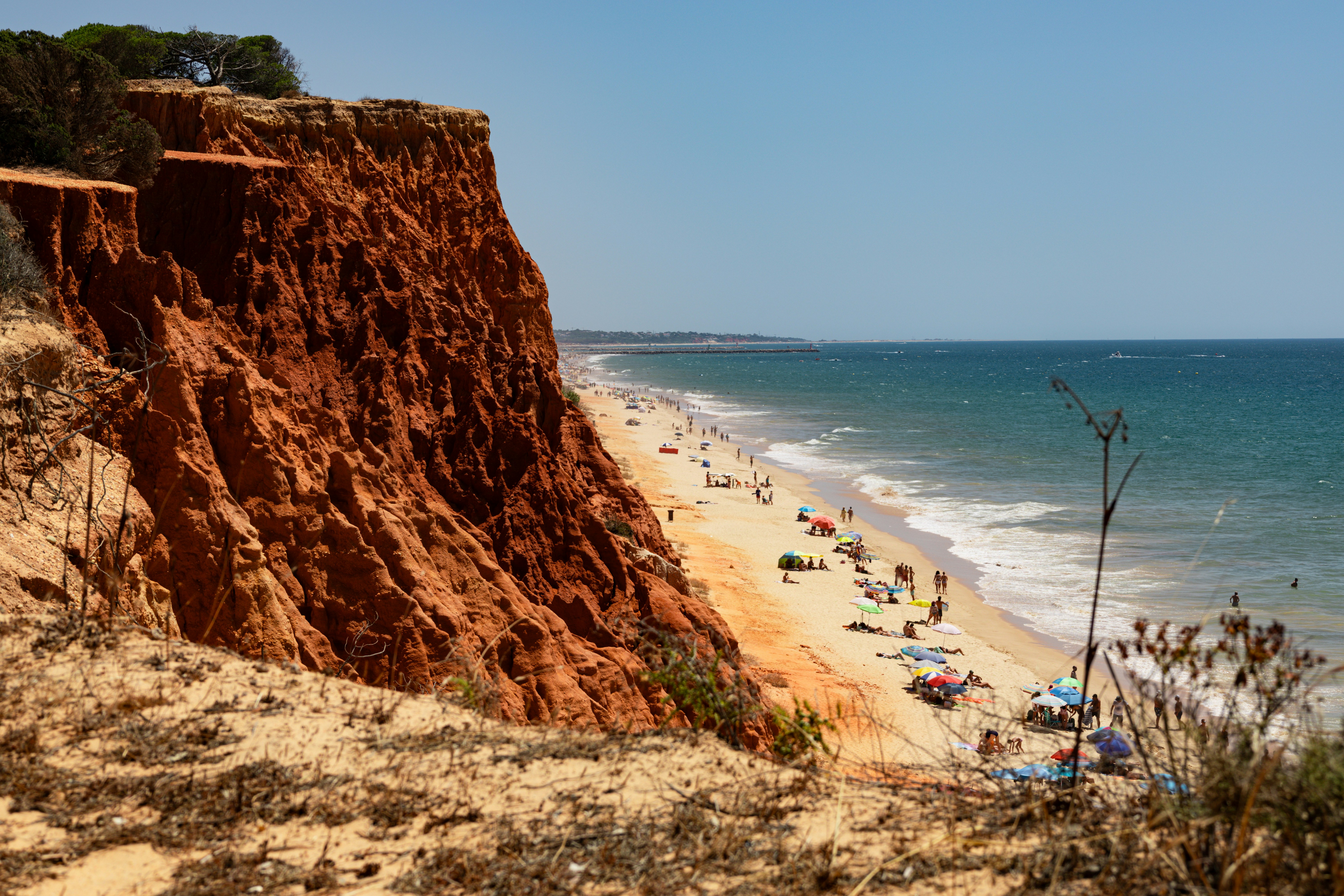 Steep red cliffs rise above a sunlit beach lined with colorful umbrellas and gentle waves.