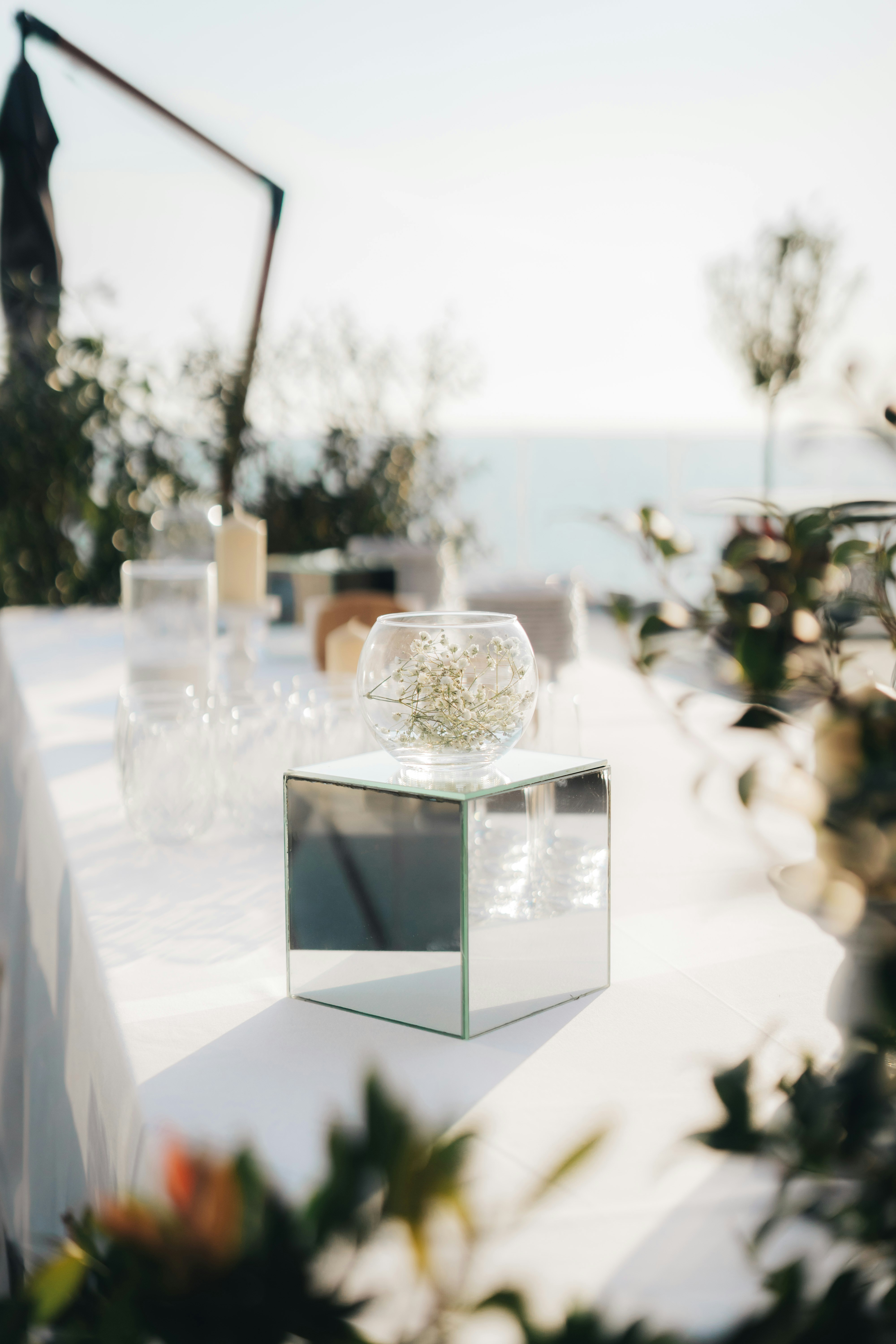 Bright seaside table setting featuring a glass cube centerpiece topped with a glass bowl of delicate white blooms, with a softly blurred ocean backdrop.