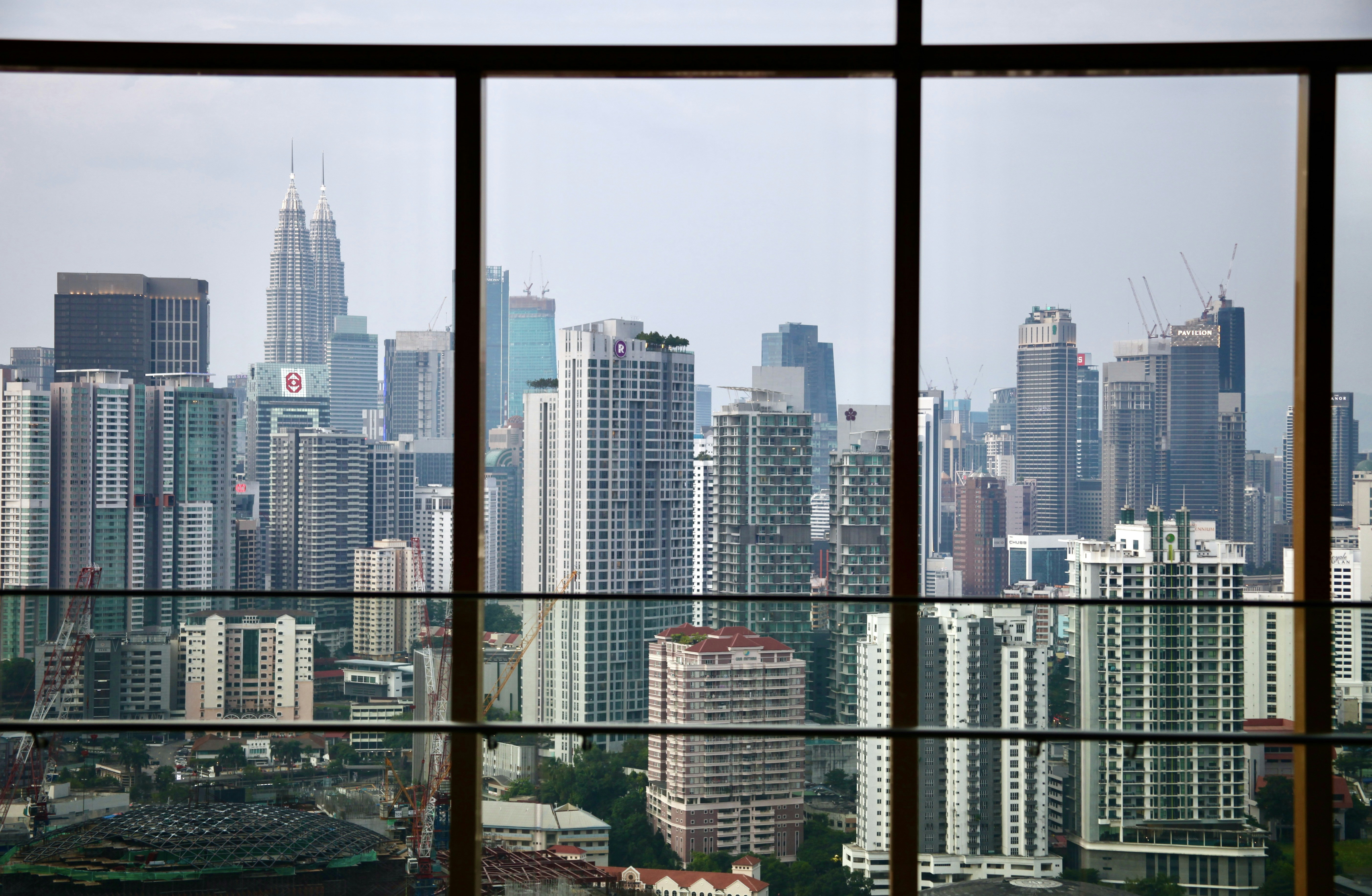 City skyline viewed through a large glass window, showcasing a blend of modern architecture and natural elements. The scene captures the vibrant life of an urban landscape.