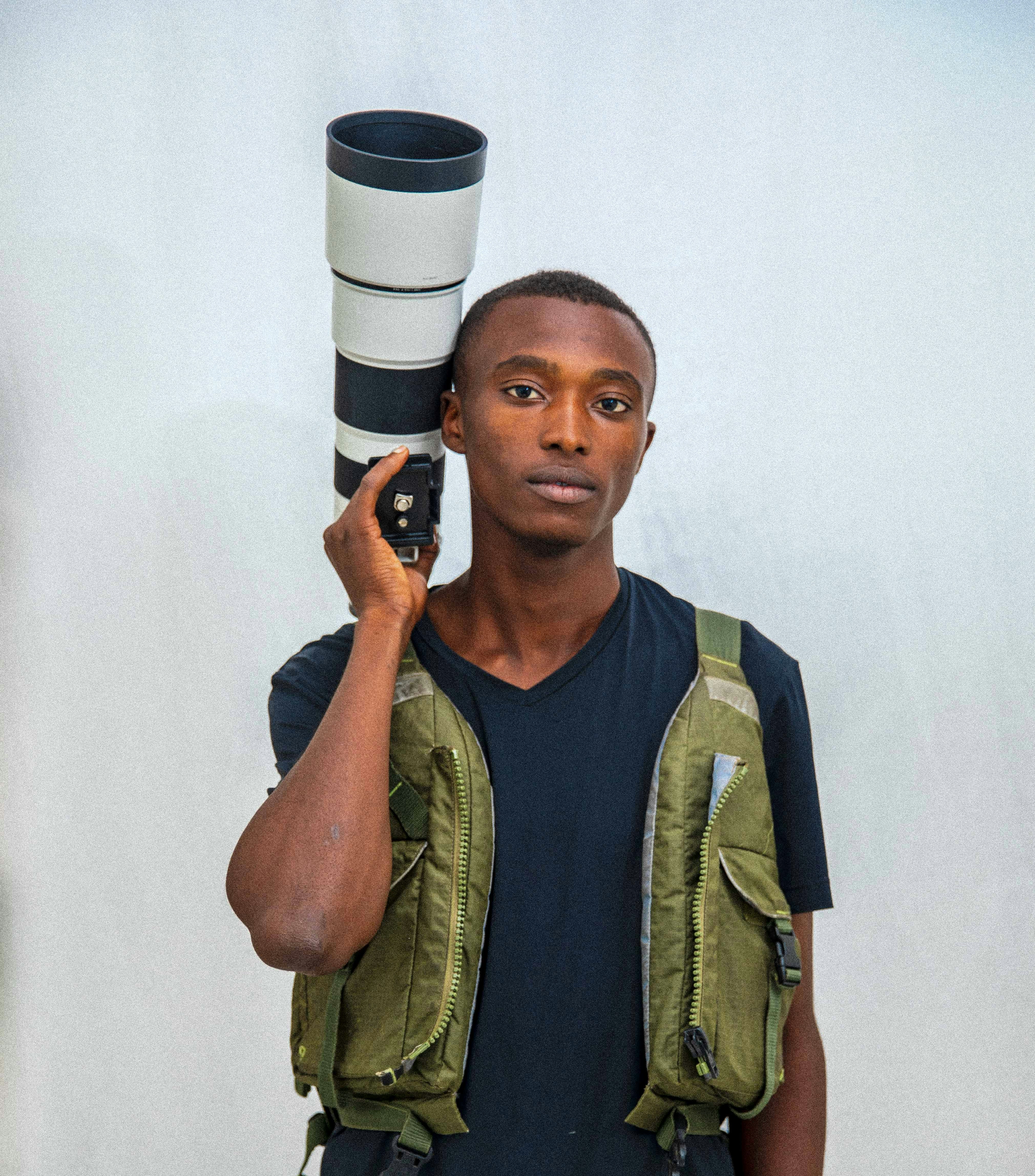 Person holding a large telephoto lens over shoulder against a plain background.