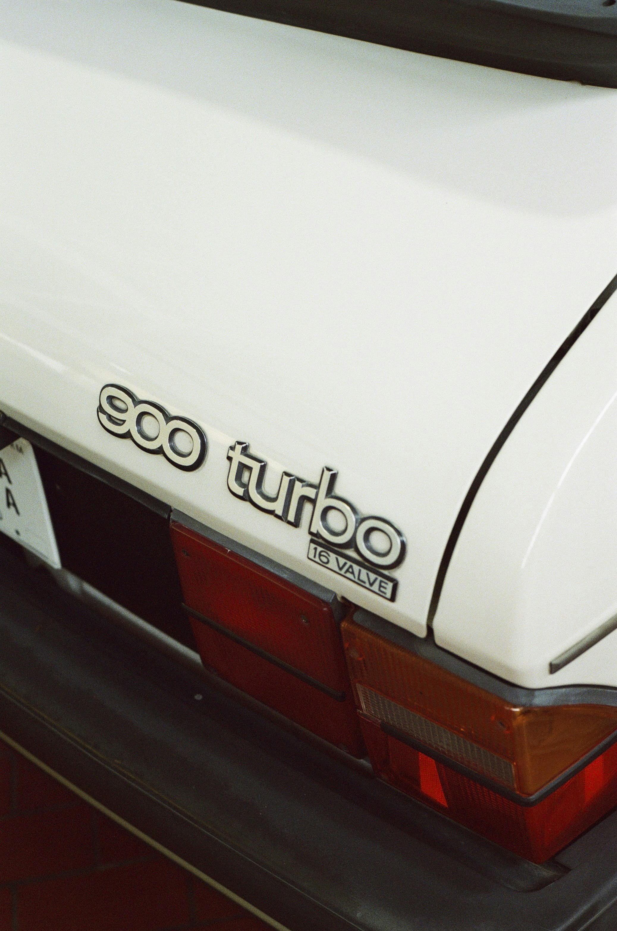 Close-up of the Saab 900 Turbo 16 Valve rear badge on a white trunk, with red taillights and a black bumper visible.