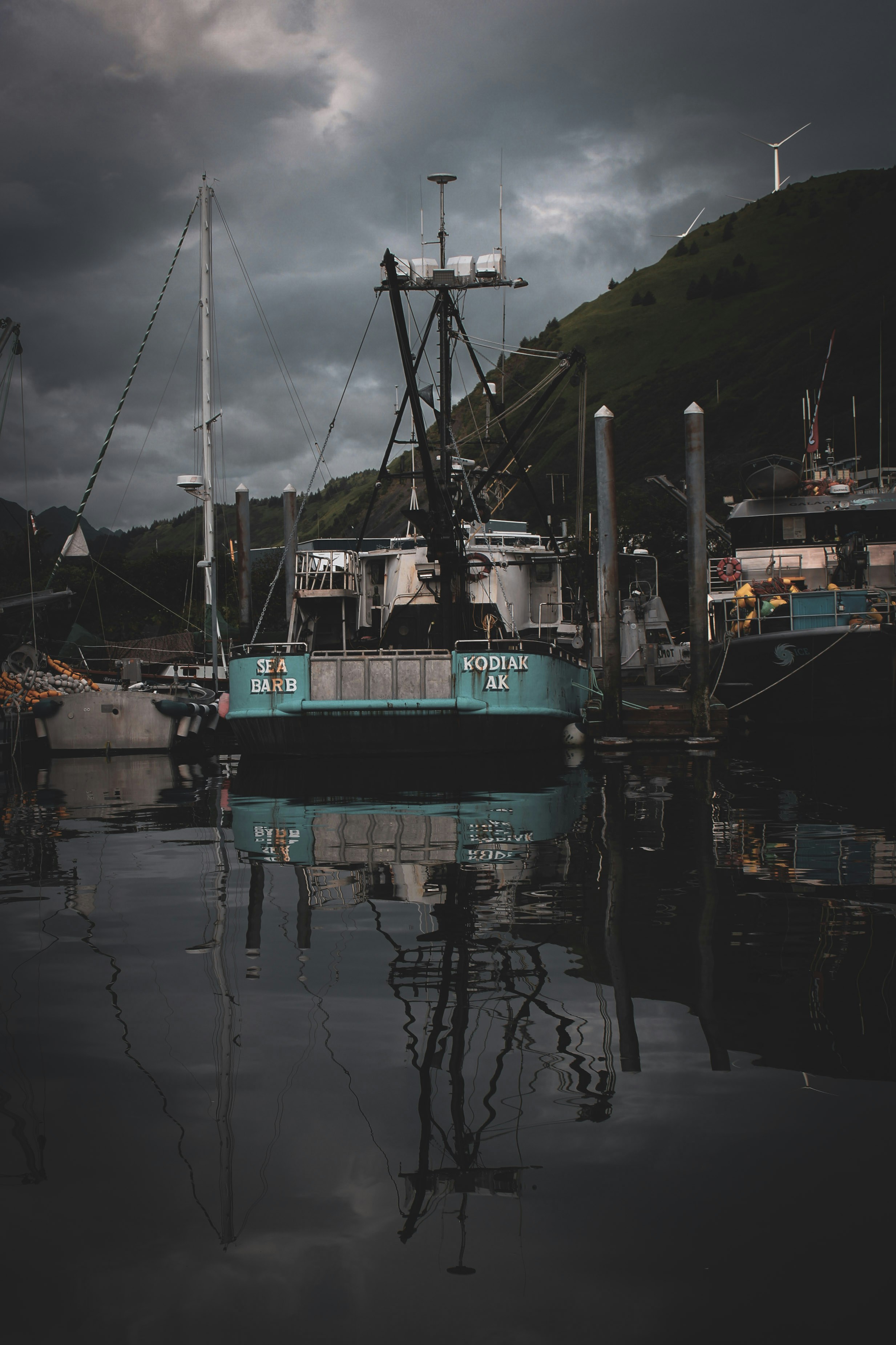 Fishing boats moored in a tranquil harbor, their reflections shimmering on the dark water under a brooding sky.