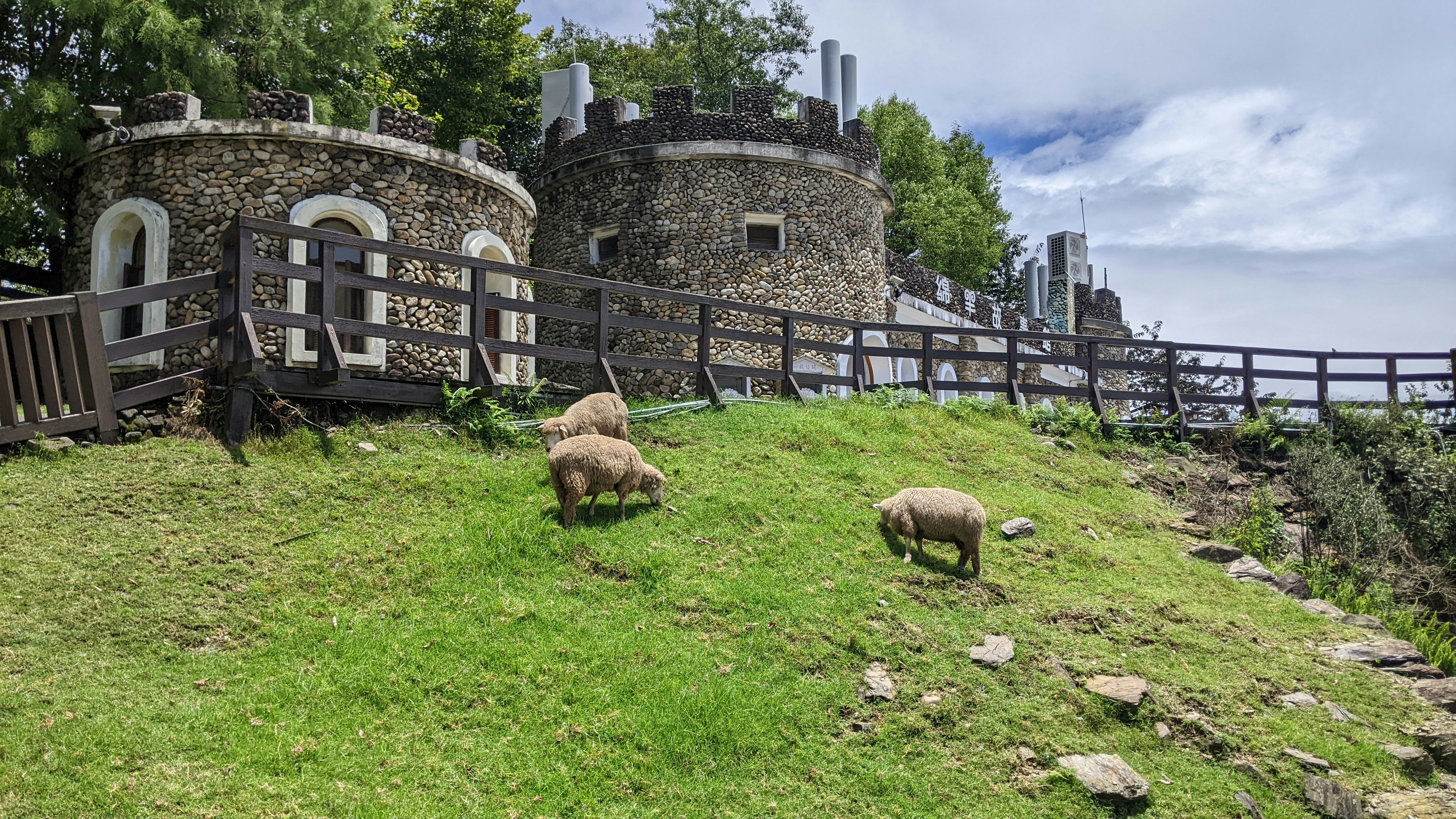 Sheep grazing on a grassy hill near a rustic stone structure under a partly cloudy sky.