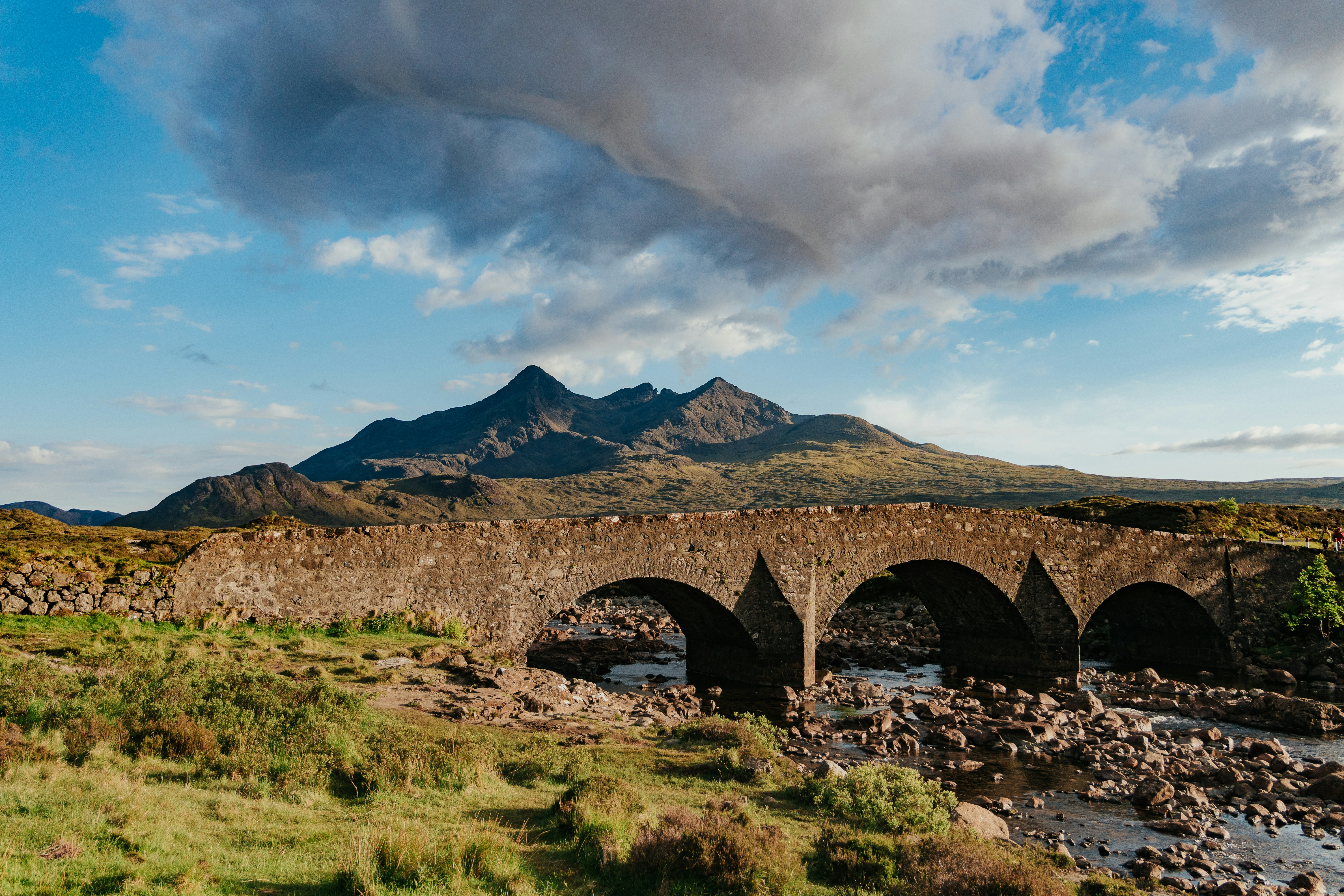 Stone arch bridge spanning a rocky river with mountains in the background under a partly cloudy sky.