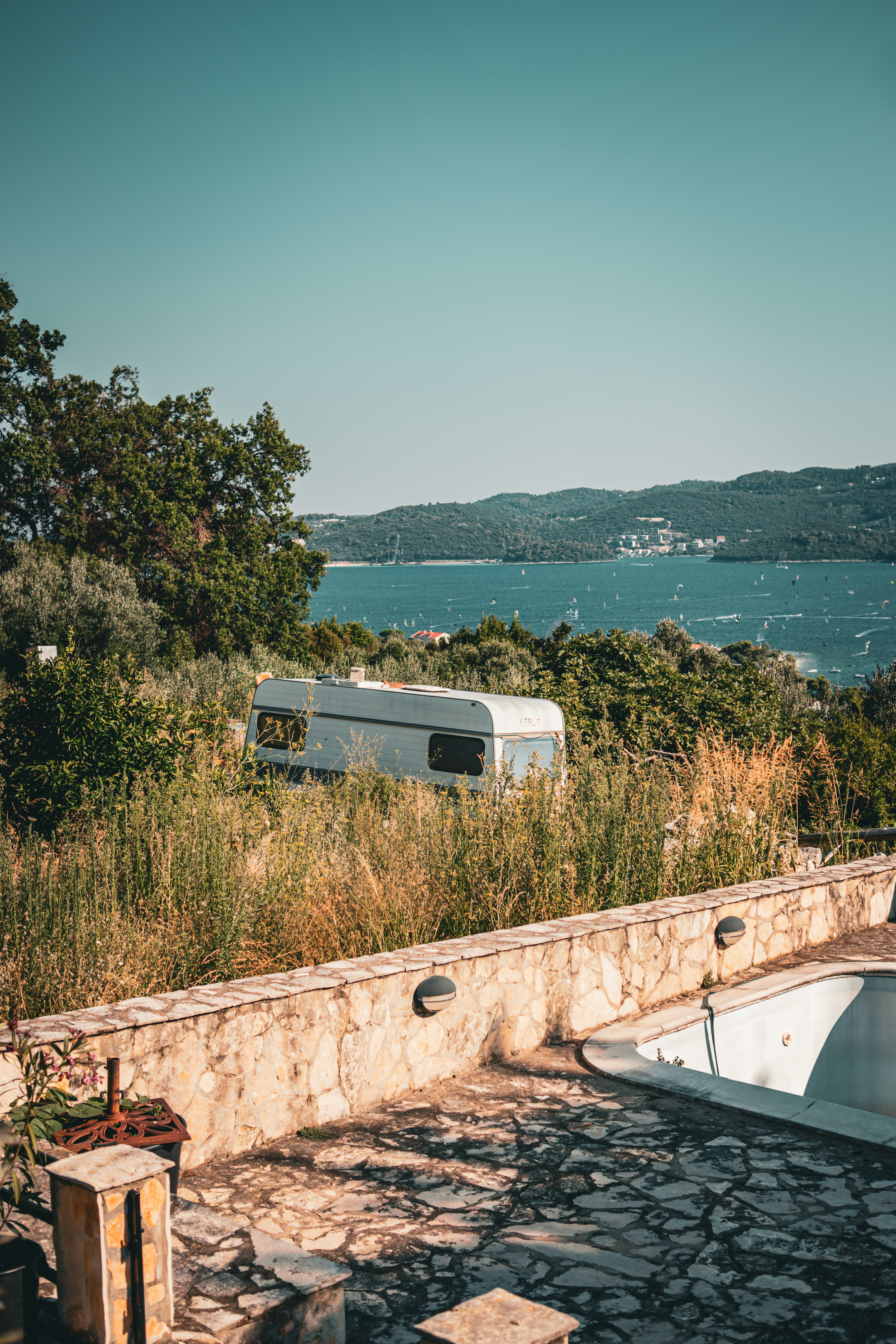 A vintage caravan nestled among overgrown vegetation, overlooking a tranquil bay dotted with sailboats. The scene captures a blend of nature and nostalgia.