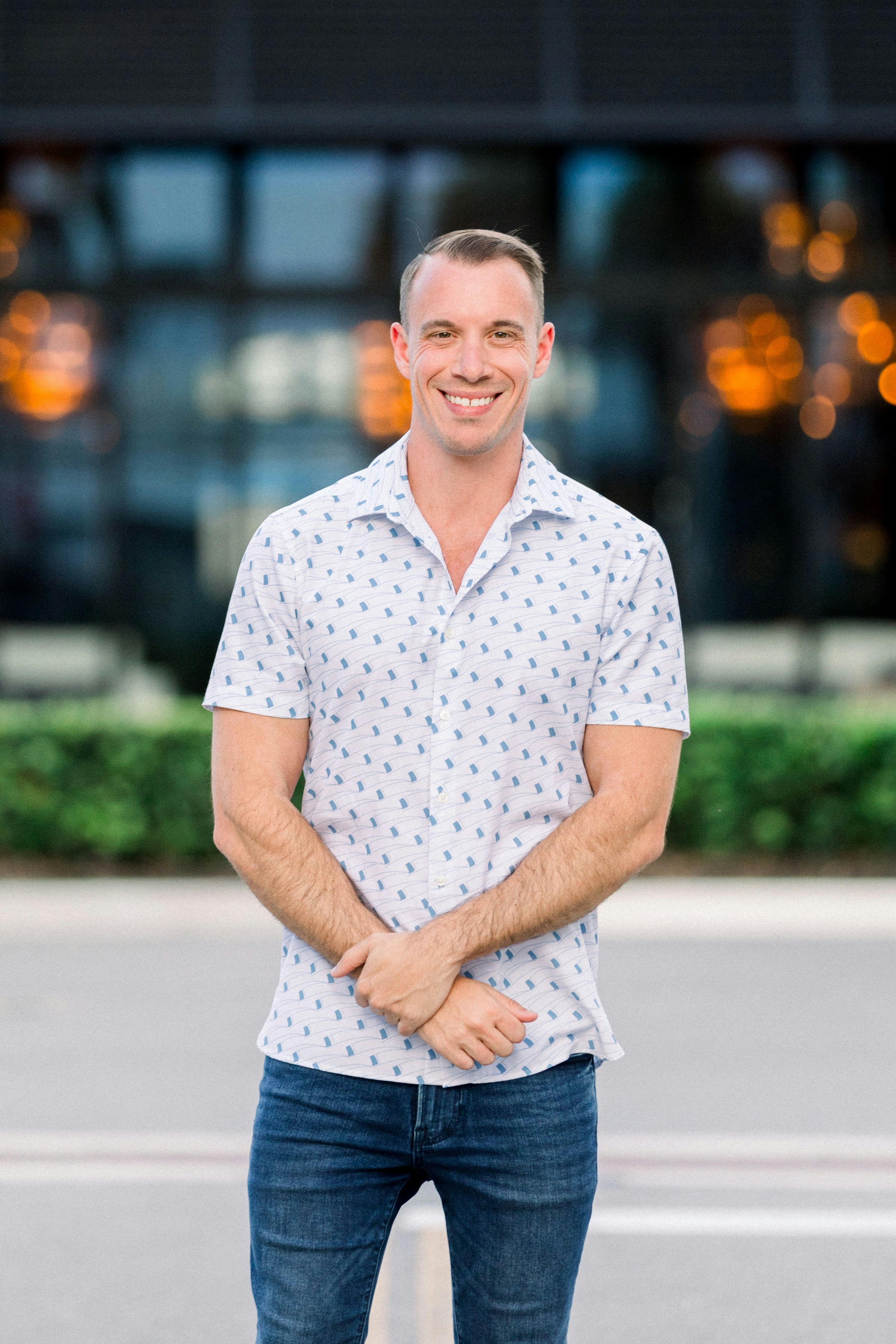 Smiling man in a patterned shirt stands confidently outdoors, framed by a softly blurred background of warm lights and greenery.