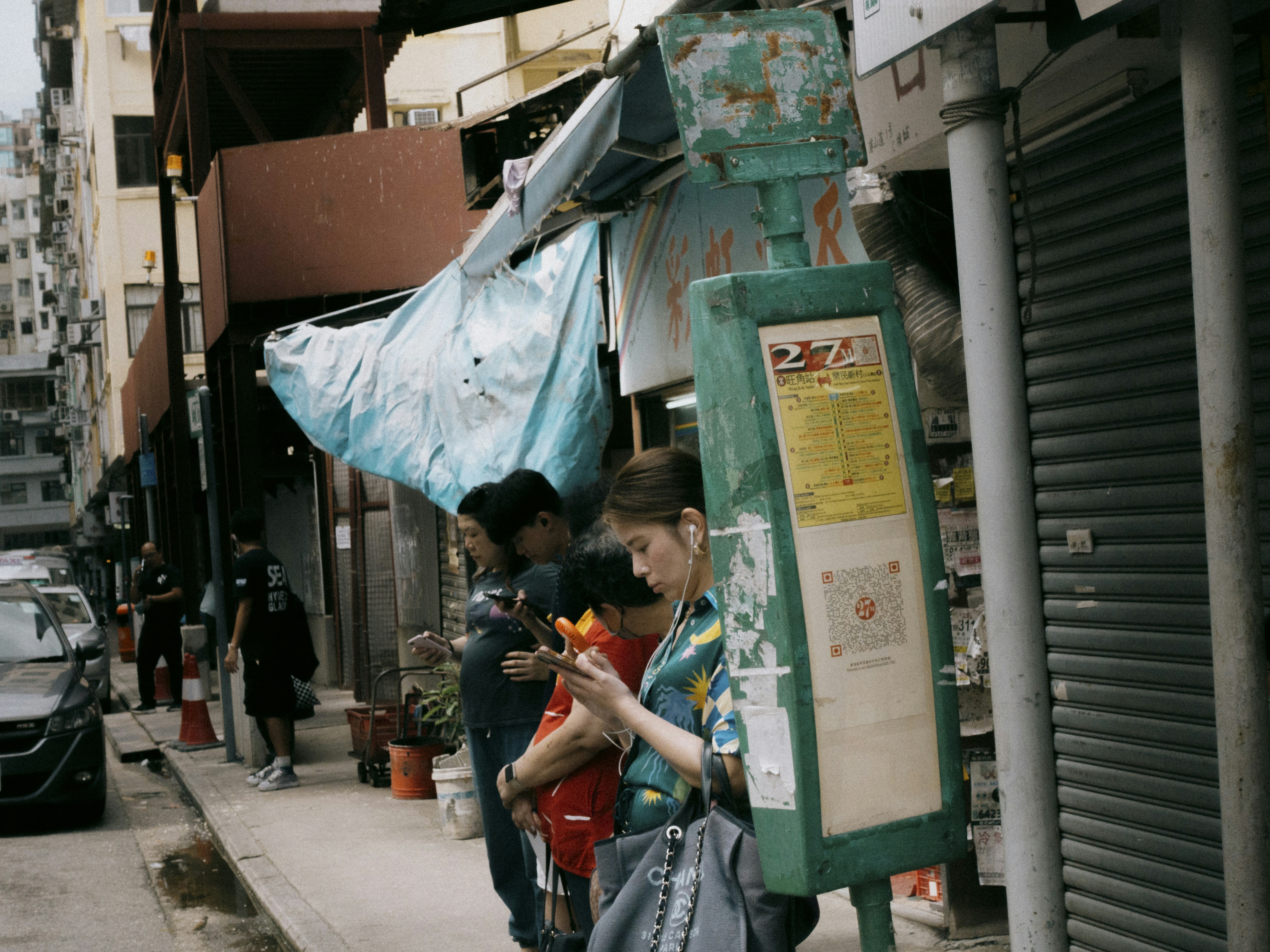 Quiet Focus at the Curb: A Woman Checks Her Phone Under a Weathered Blue Canopy