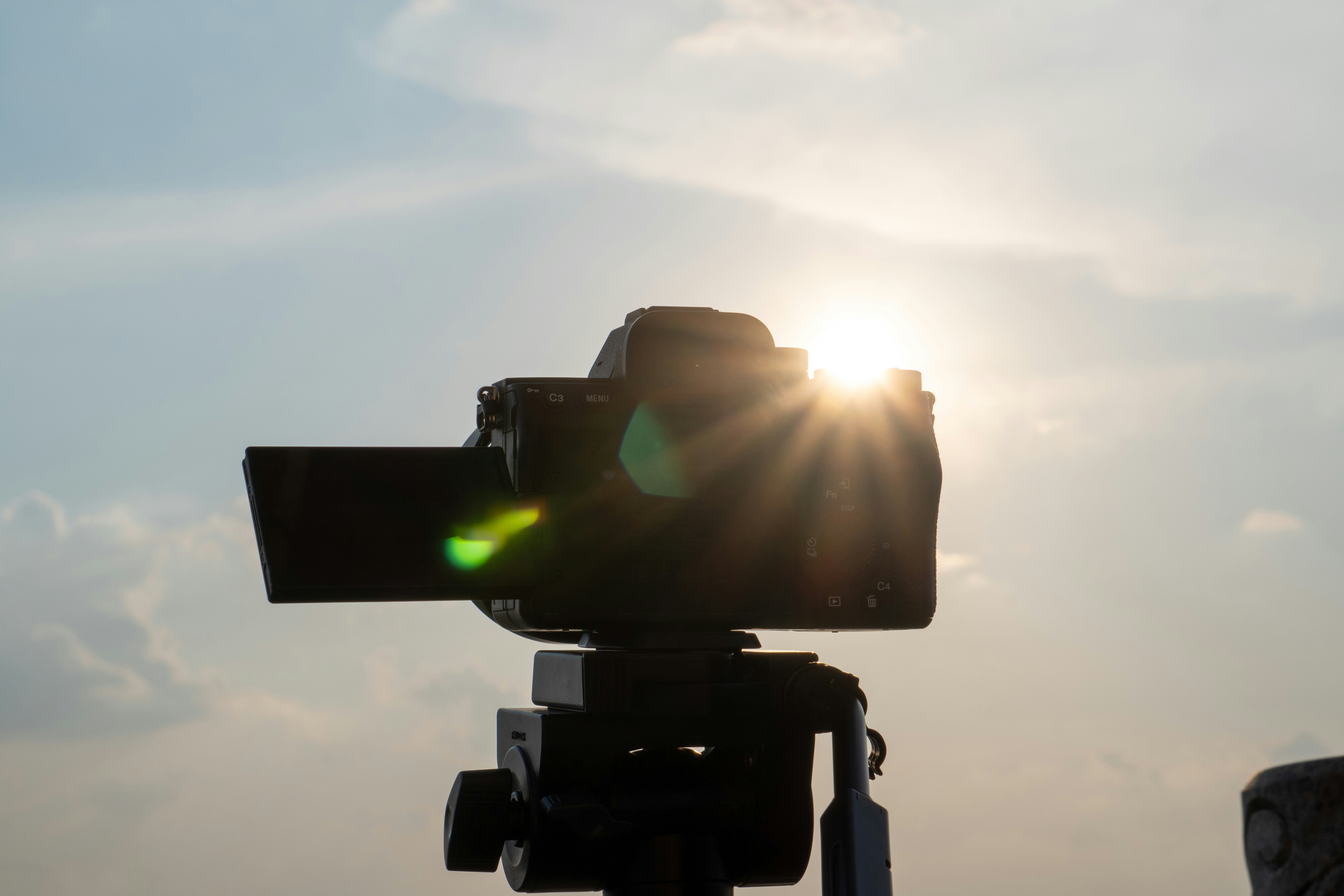 This striking image features a camera silhouetted against the setting sun, creating a captivating play of light and shadow. The warm glow of the sun peeks from behind the camera, casting a radiant halo and highlighting the soft blues and grays of the sky. The composition emphasizes the blend of technology and nature, making the photograph both visually intriguing and atmospheric.