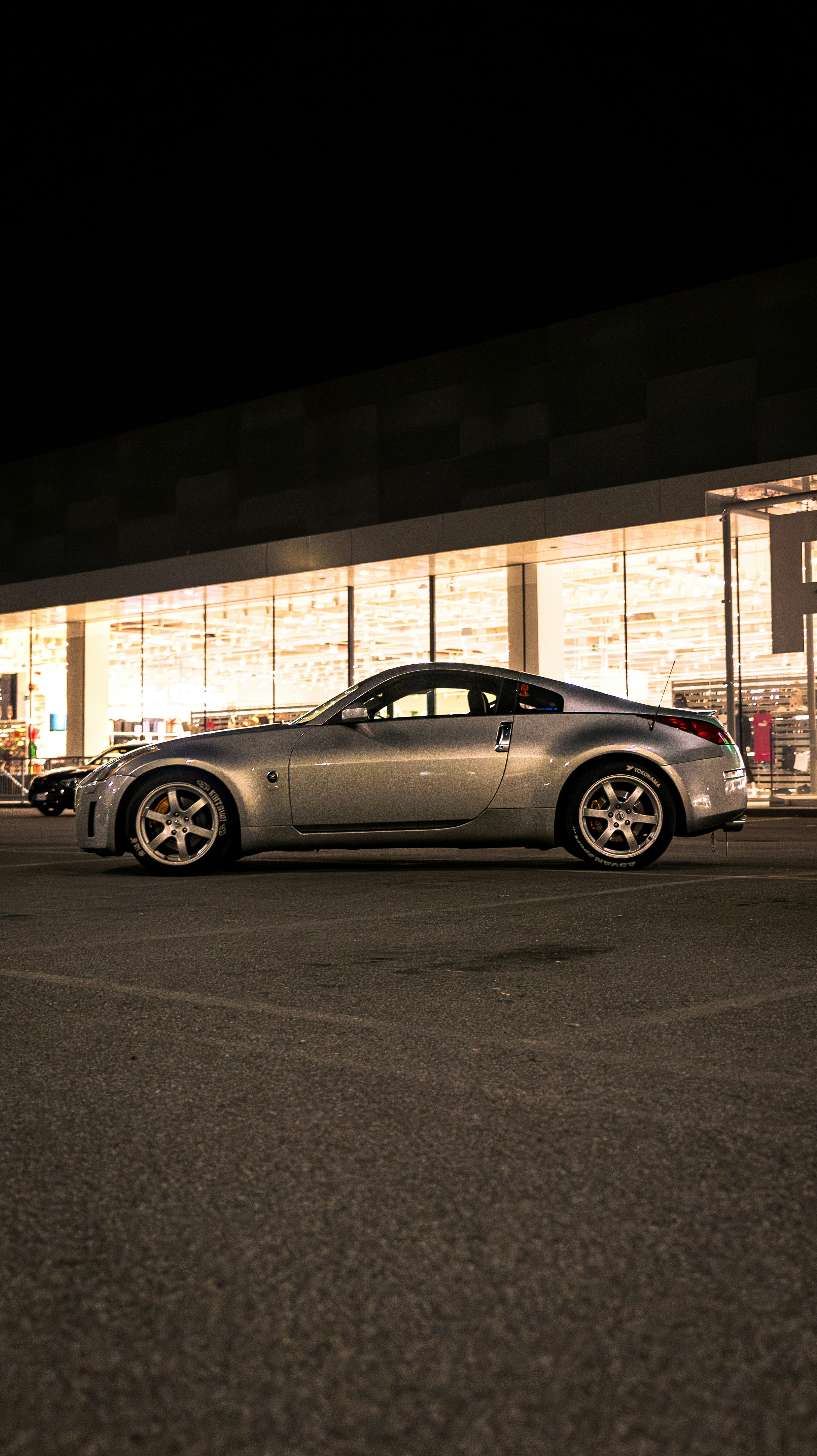 Sleek silver sports car parked in a well-lit retail area at night, showcasing its curves and design against a vibrant backdrop of illuminated storefronts.