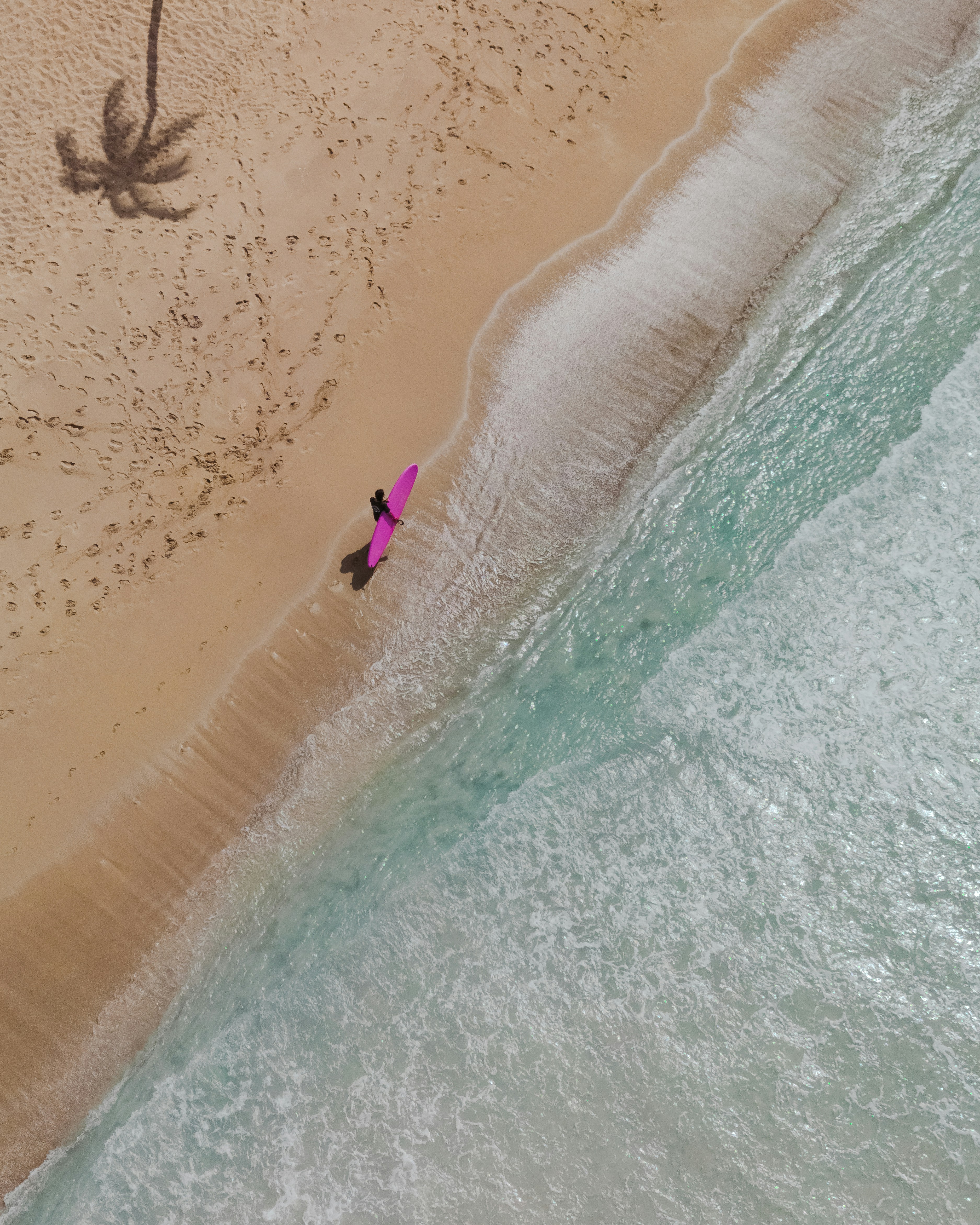 Aerial drone photograph of a sandy beach with turquoise water meeting the shore, featuring a pink surfboard and a lone surfer near the waterline.