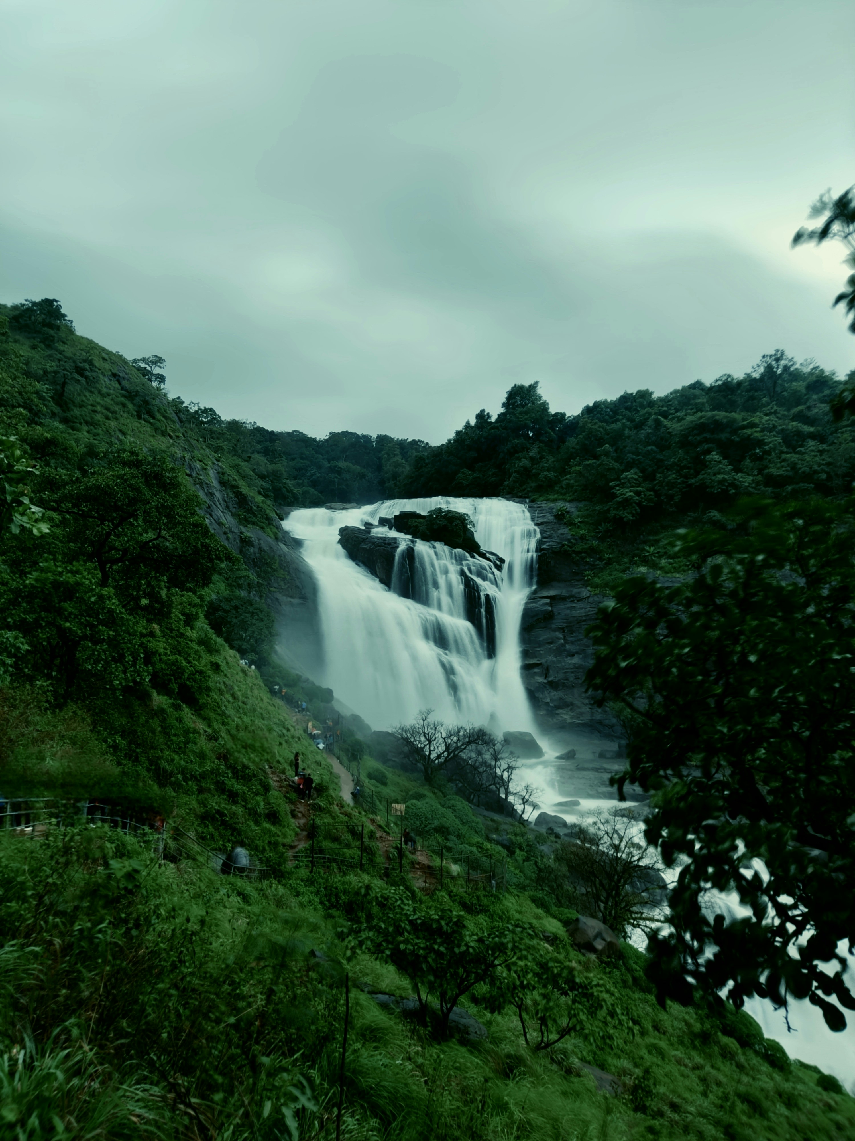 Lush valley greenery frames a multi-tier waterfall tumbling over mossy rocks beneath a muted, overcast sky.