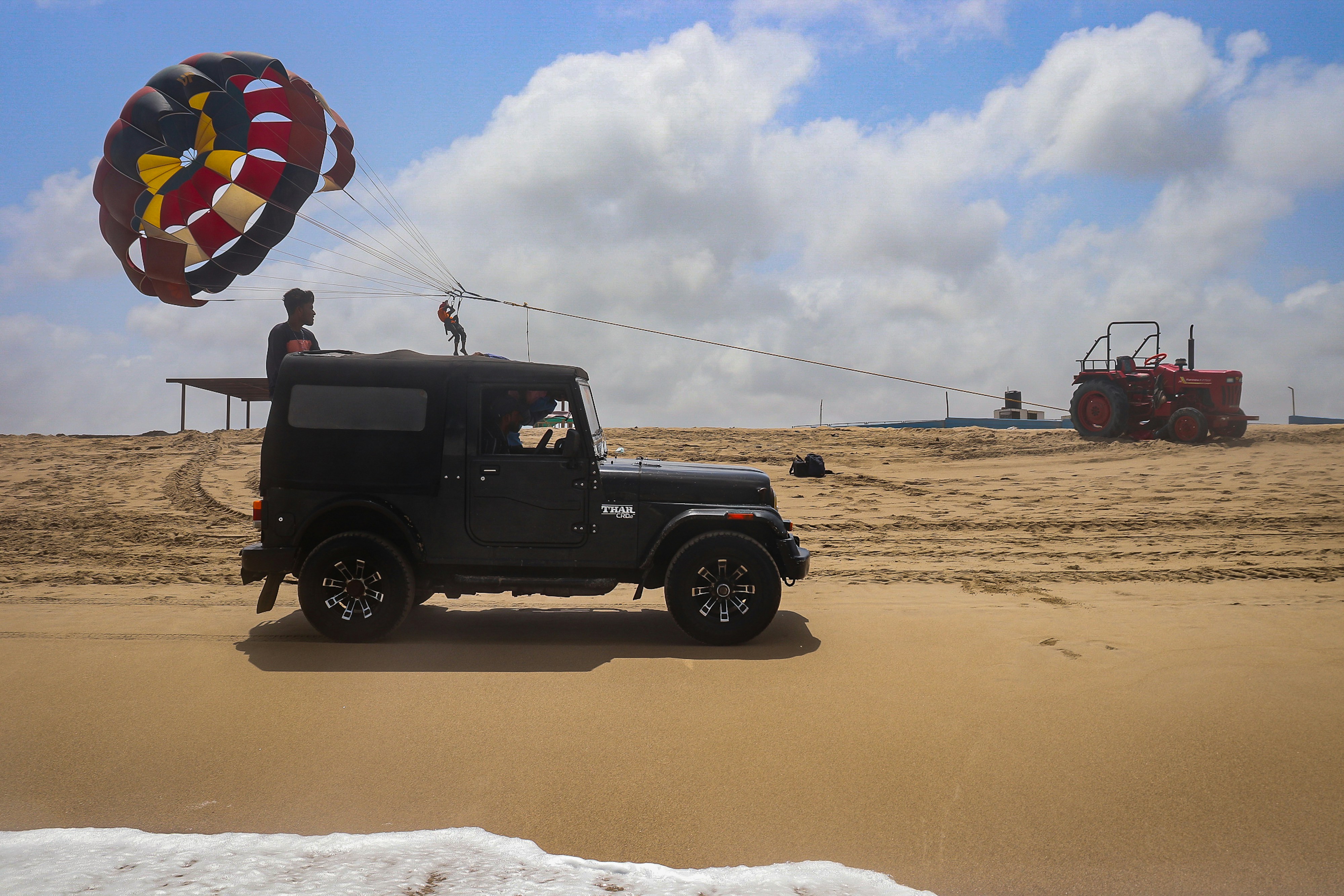 Parachutist lands near a parked jeep on a sandy beach, with a red utility vehicle in the background. The scene captures action on a bright, partly cloudy day with tire tracks in the sand.