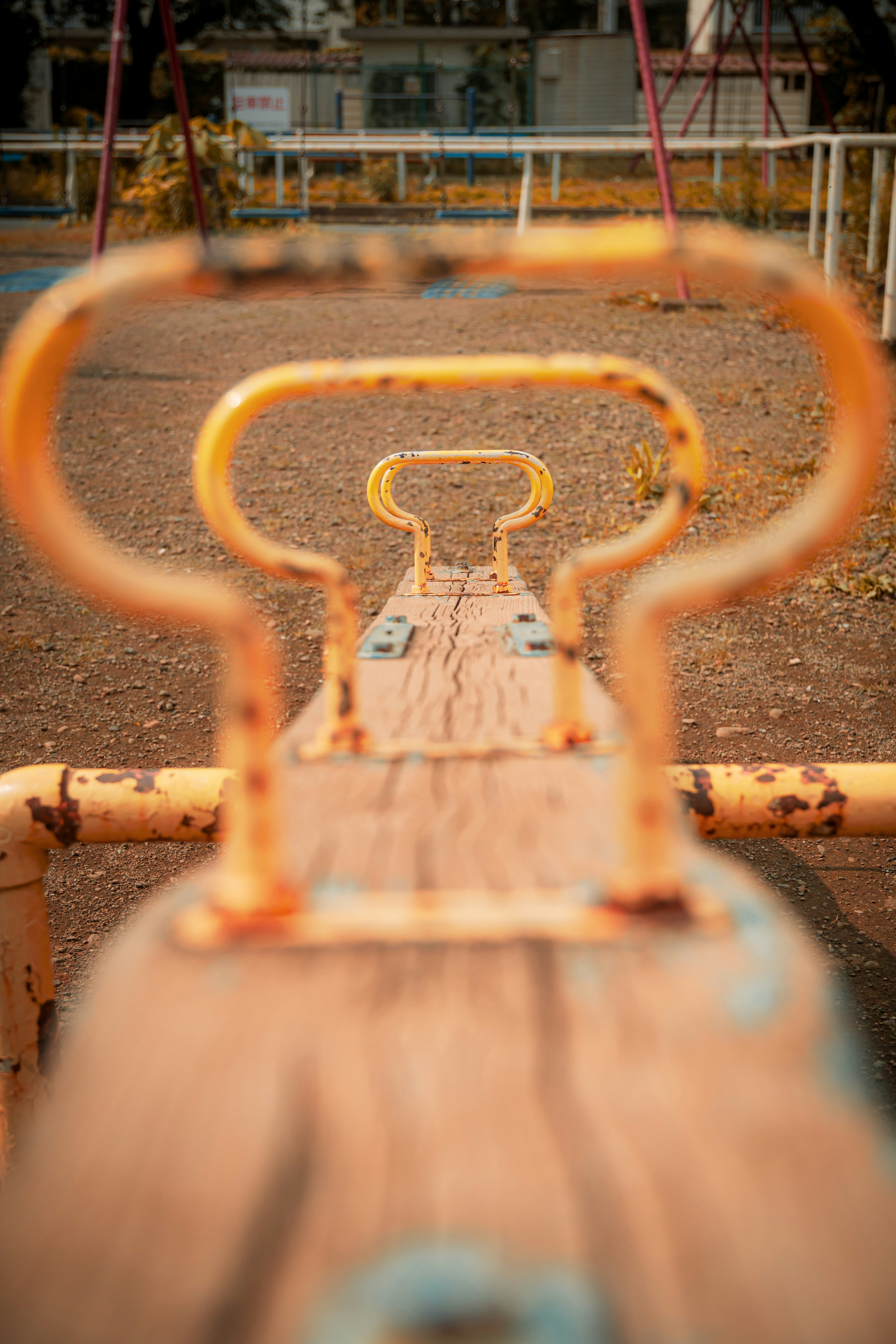 A weathered seesaw's perspective, showcasing the vibrant yellow handles leading into the distance. The ground is scattered with autumn leaves, adding a nostalgic touch.