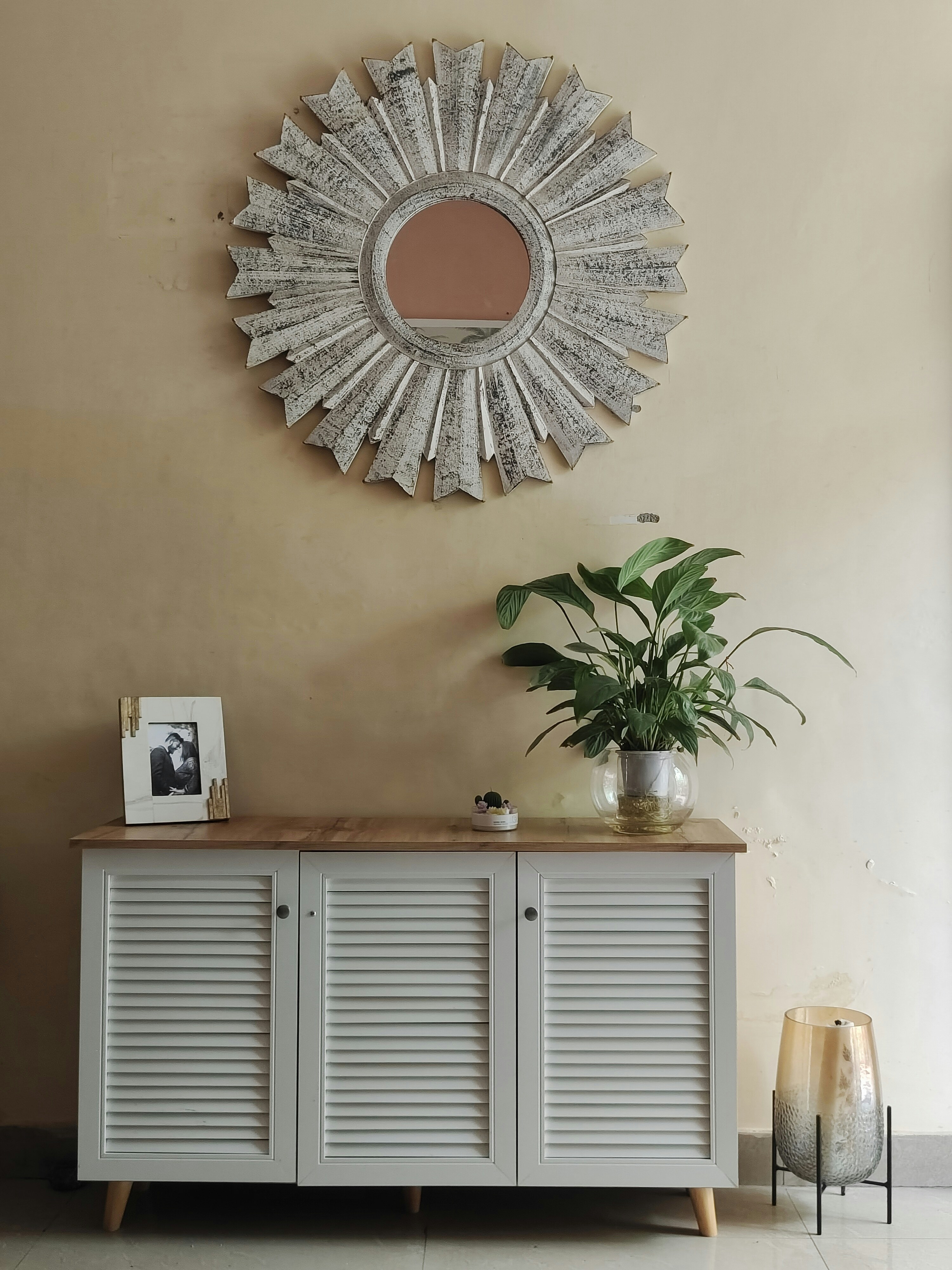 Photograph of a sunburst mirror above a white louvered cabinet with a potted plant, photo frame, and glass vessel in a softly lit beige room.