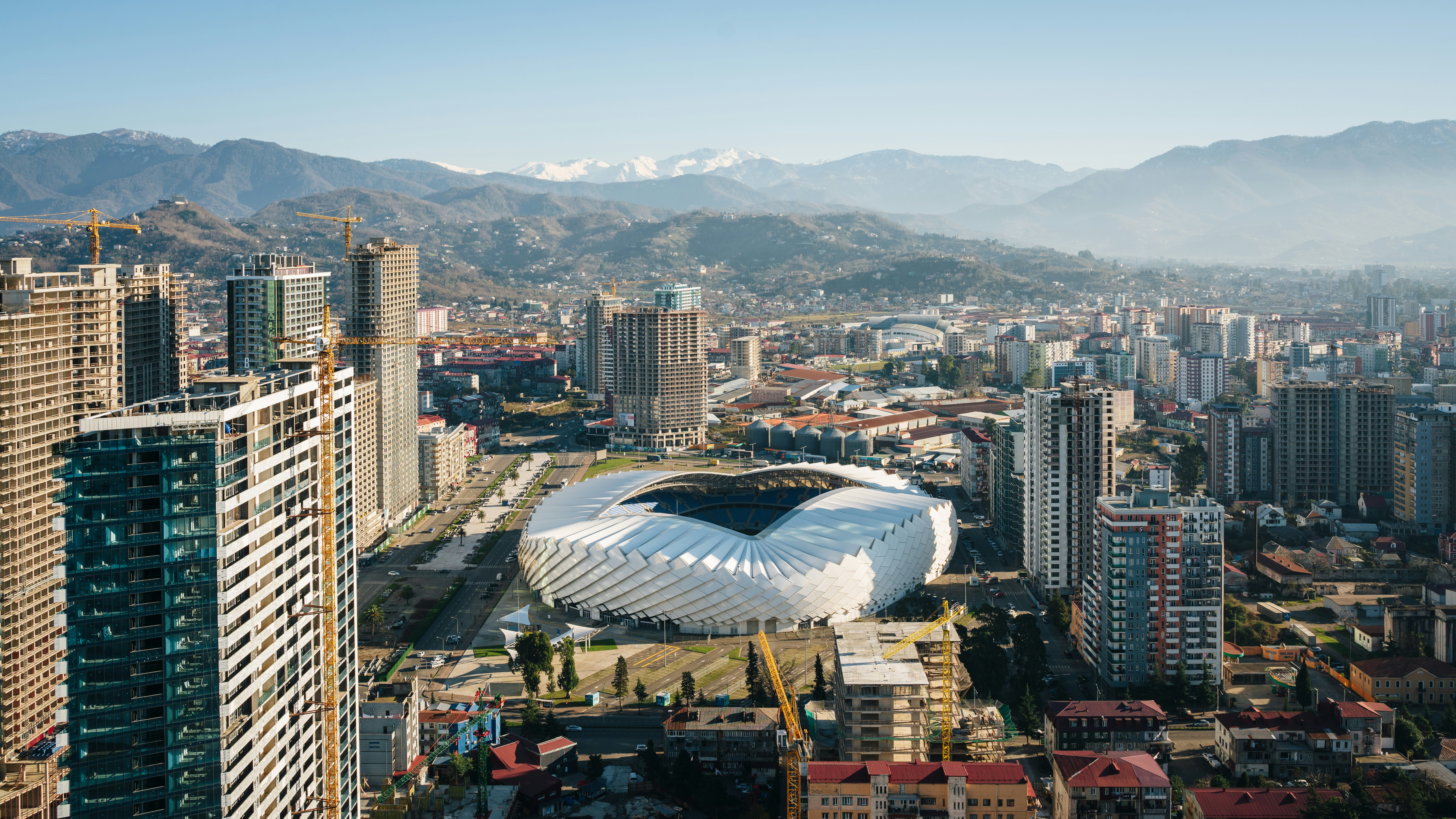 Aerial view of a modern stadium surrounded by urban architecture, showcasing the blend of sports and city life. Snow-capped mountains loom in the background.