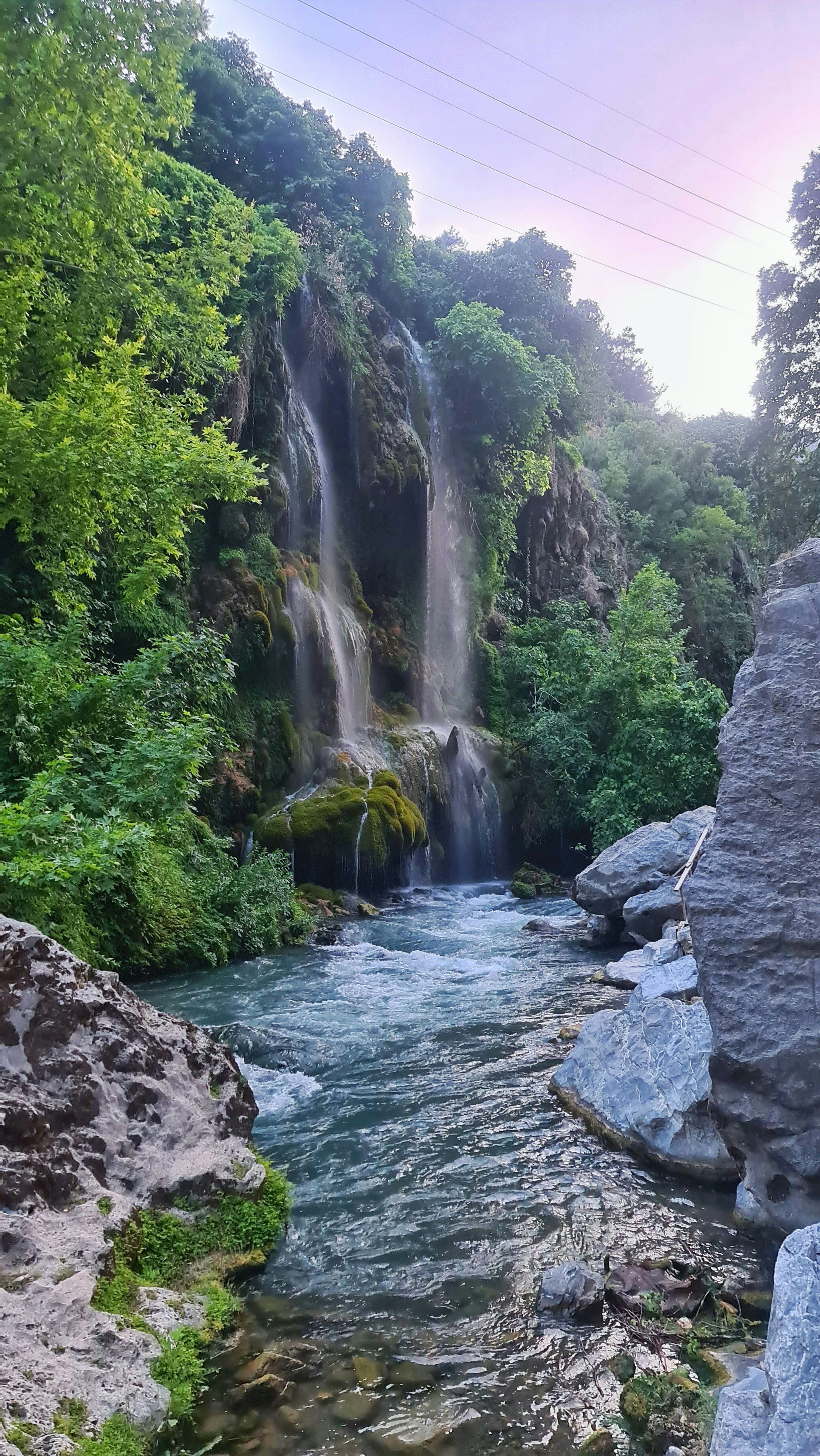 Waterfall cascading over moss-covered rocks surrounded by lush greenery, with a serene river flowing below.