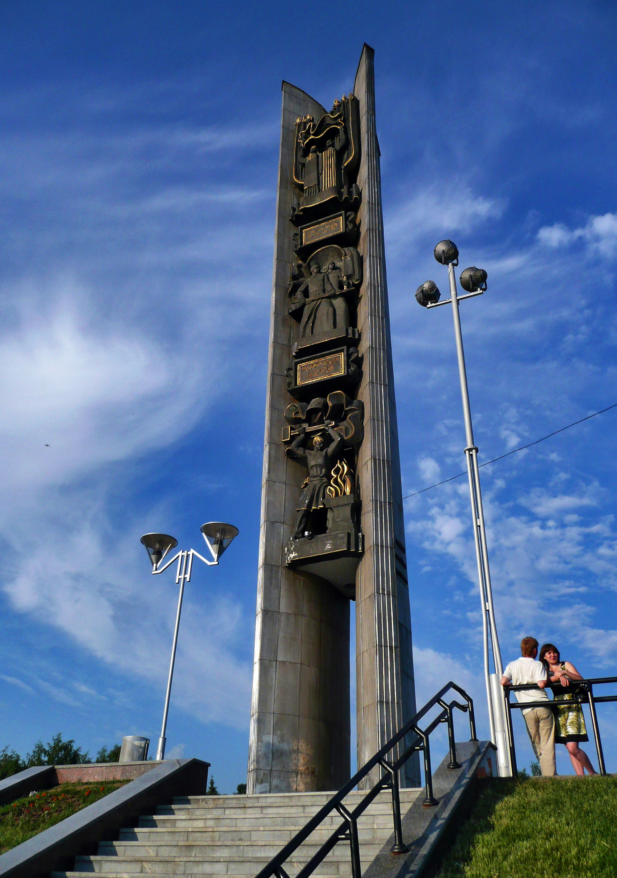 A tall monument adorned with intricate sculptures representing cultural themes, accompanied by modern street lamps and a couple enjoying the view.