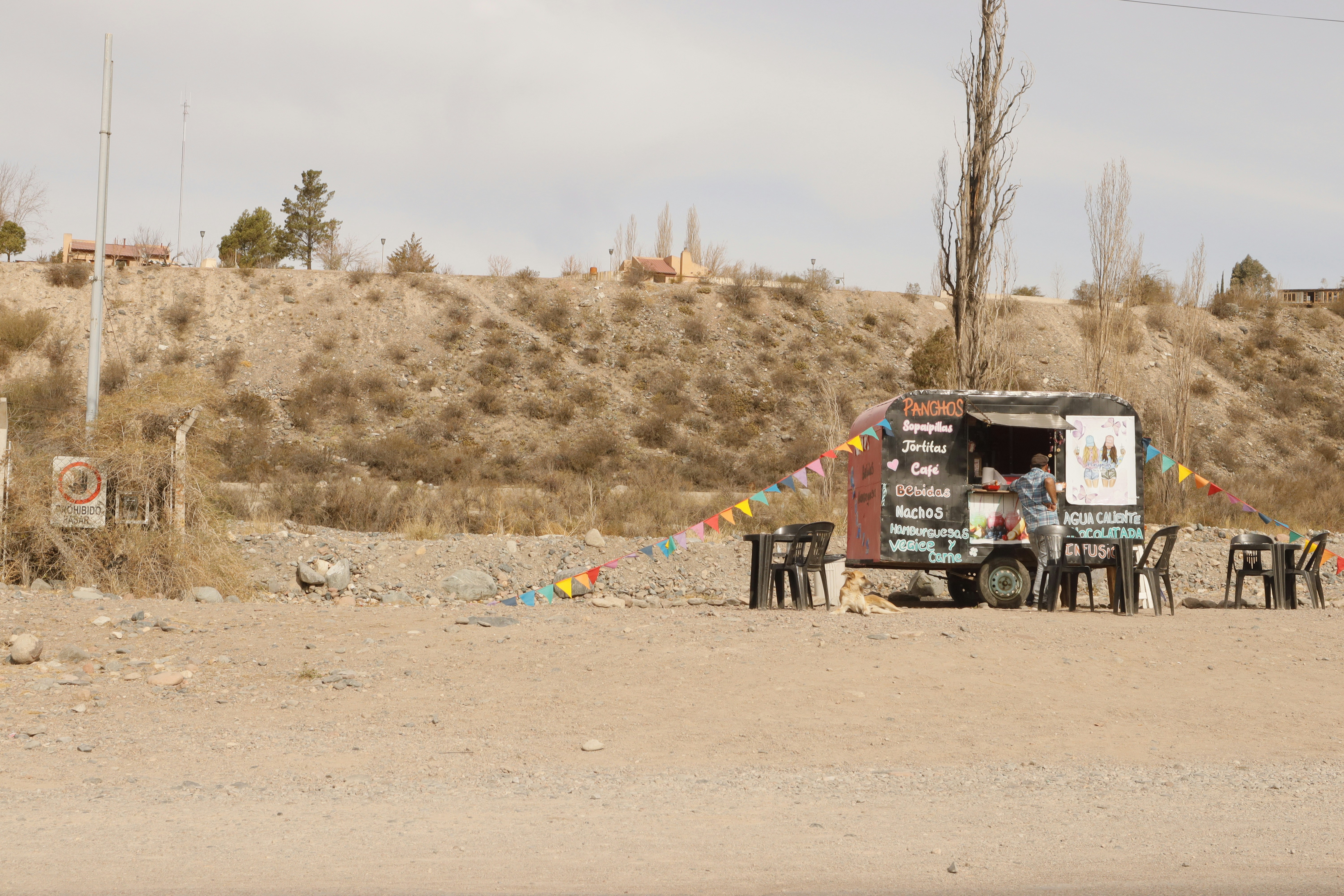 Food truck parked beside a dirt road with colorful flags fluttering in a barren landscape.