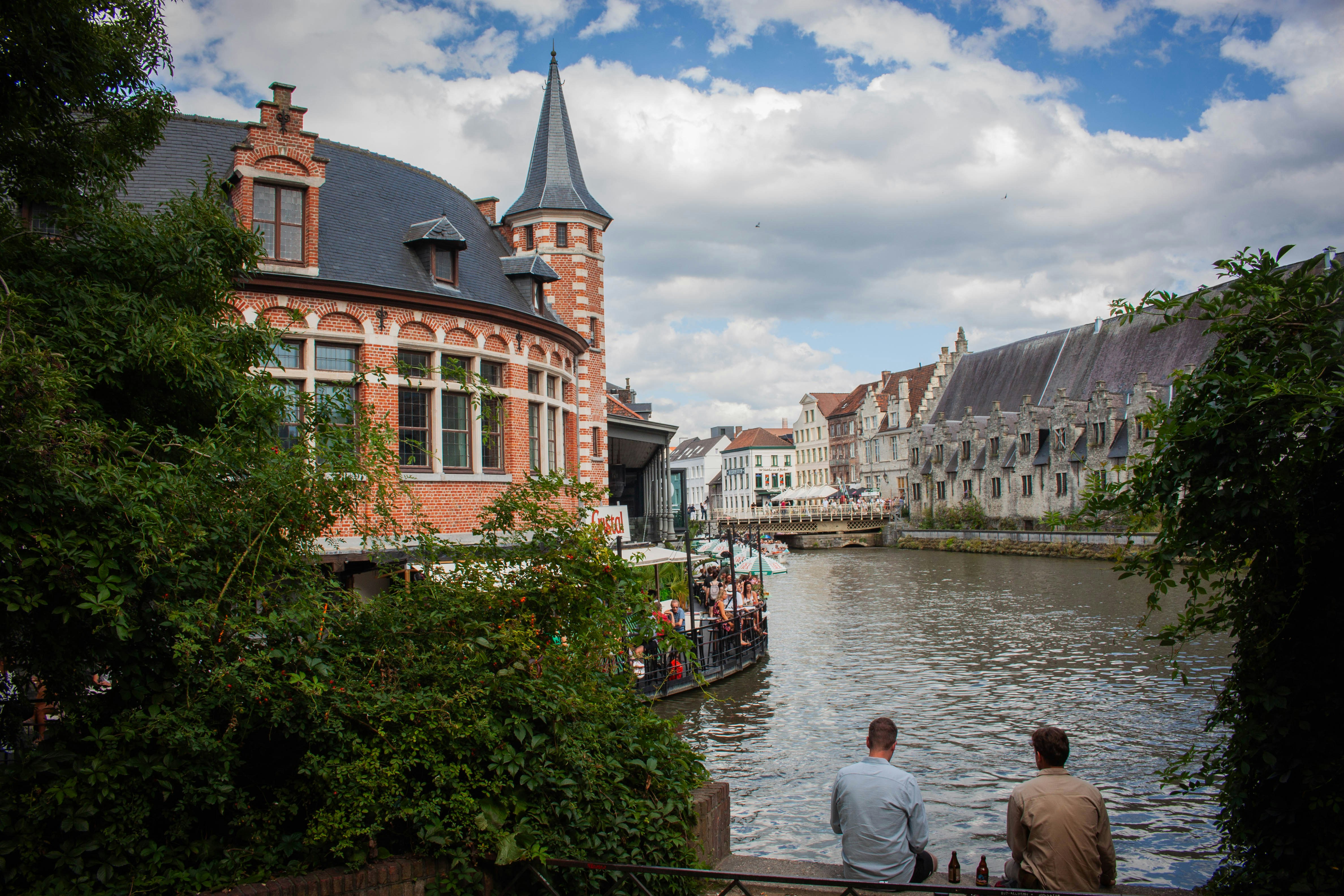Two people sit by a tranquil river, surrounded by historic architecture under a partly cloudy sky.
