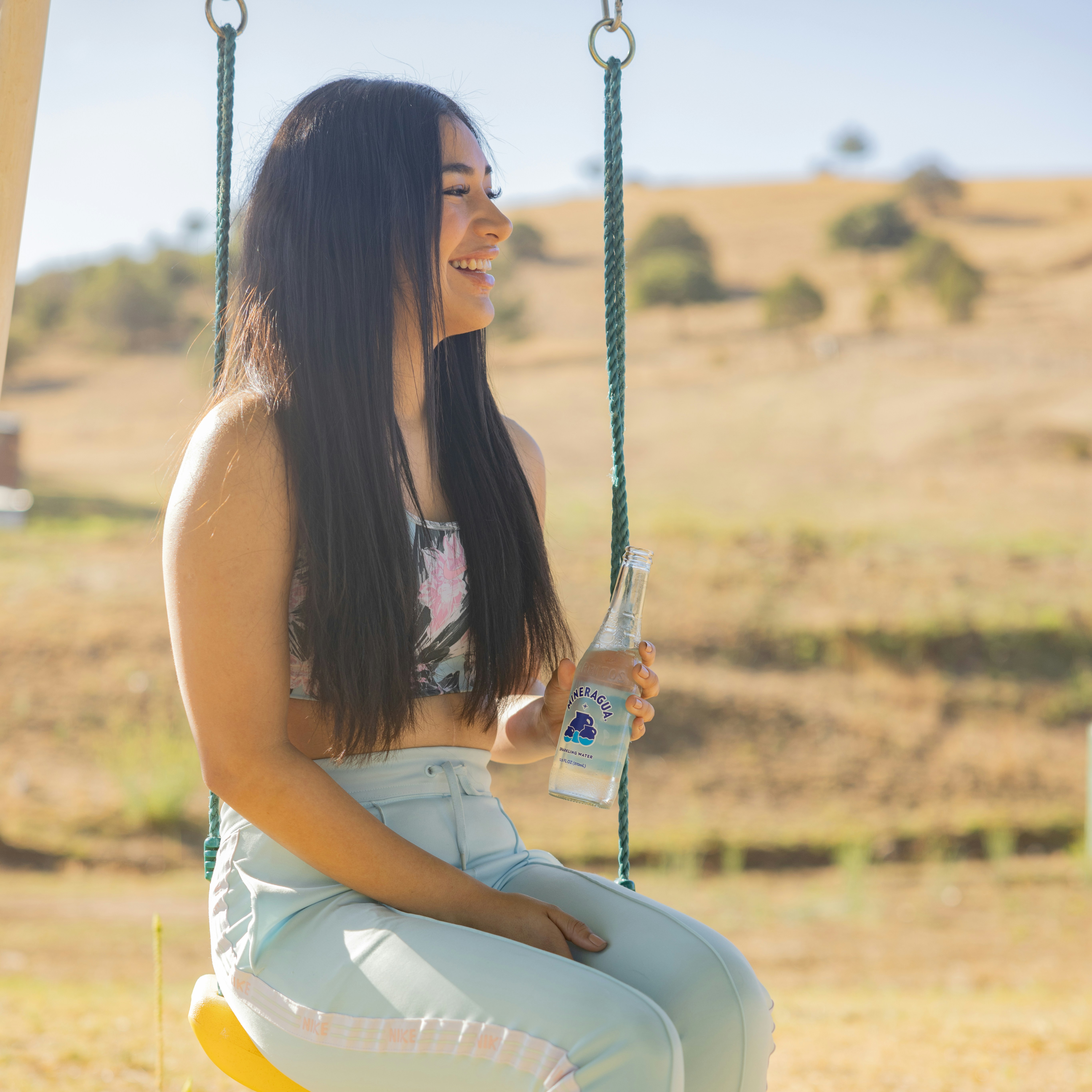 A woman with long dark hair sits on a swing in a sunlit outdoor setting, smiling while holding a bottle. Golden hills glow in the background, creating a warm, relaxed atmosphere.