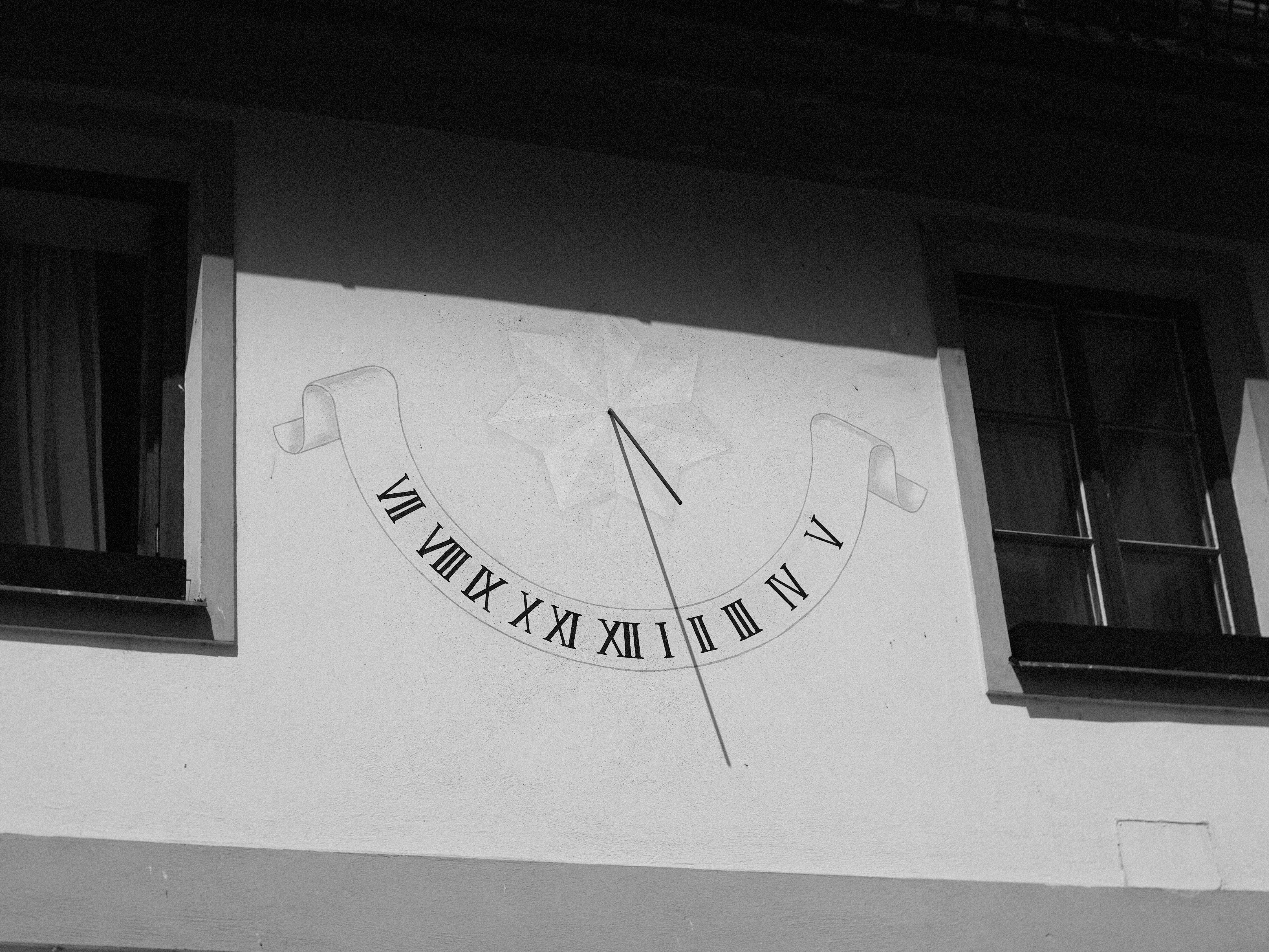 Sundial mural on a building facade, featuring Roman numerals and a central gnomon casting a shadow. The design highlights the passage of time.