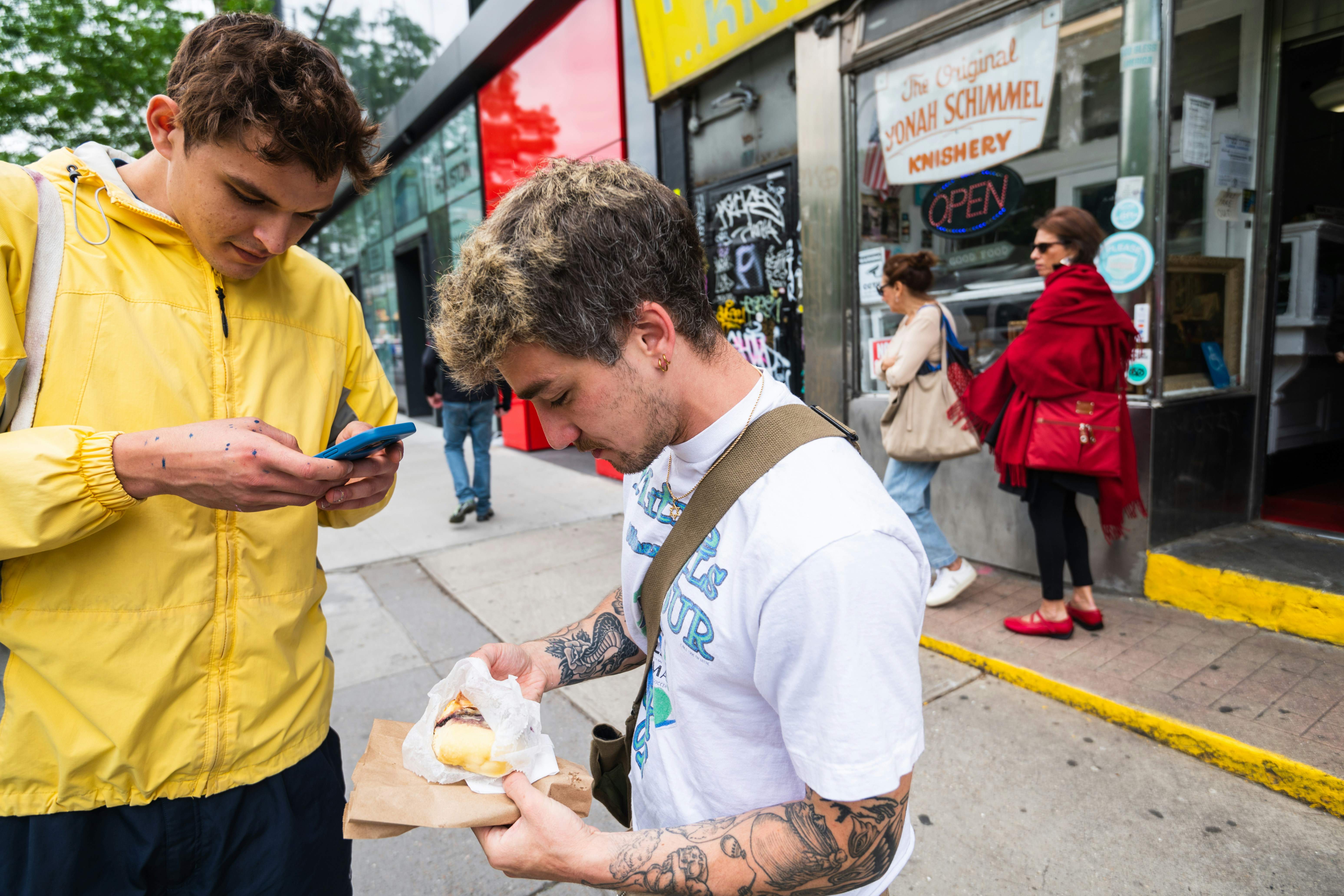 Two men stand outside a storefront, one in a yellow jacket checking his phone, the other holding a snack.