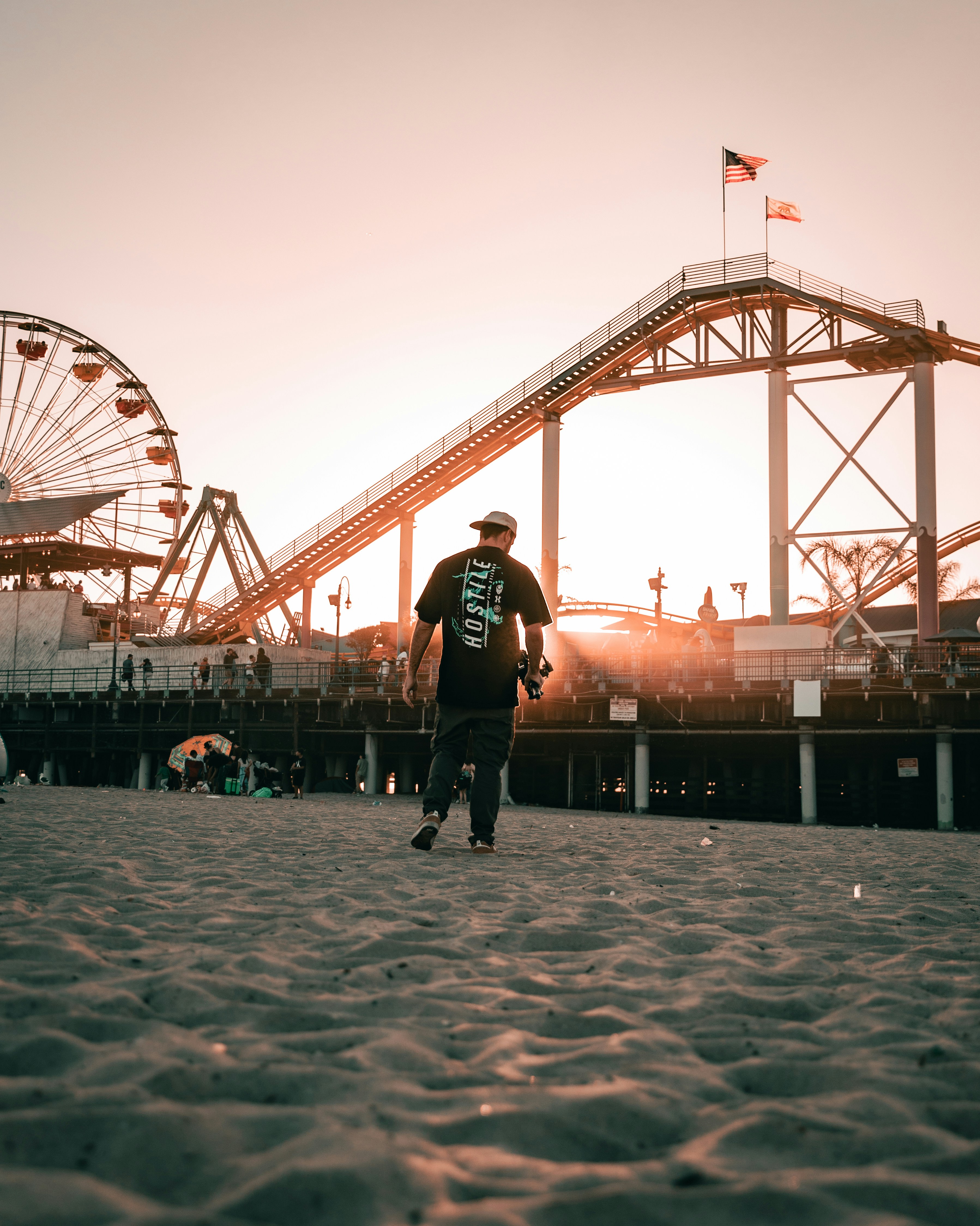 Horizon Walk at Sunset: A lone figure strides the sand under the Ferris wheel and roller coaster