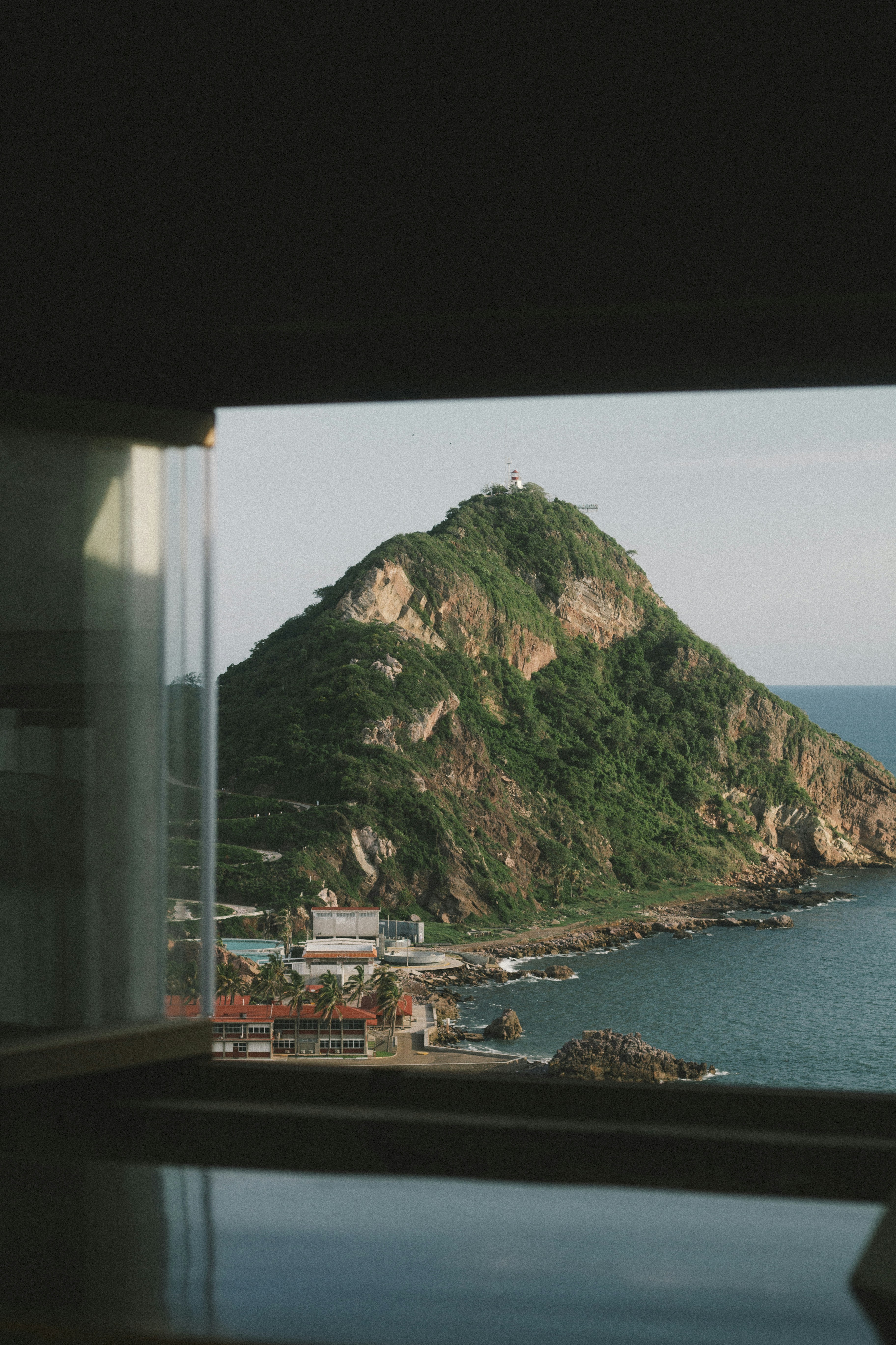 Verdant hill rising above a tranquil sea, framed by coastal architecture and a lighthouse. The scene captures the harmony between nature and human habitation.