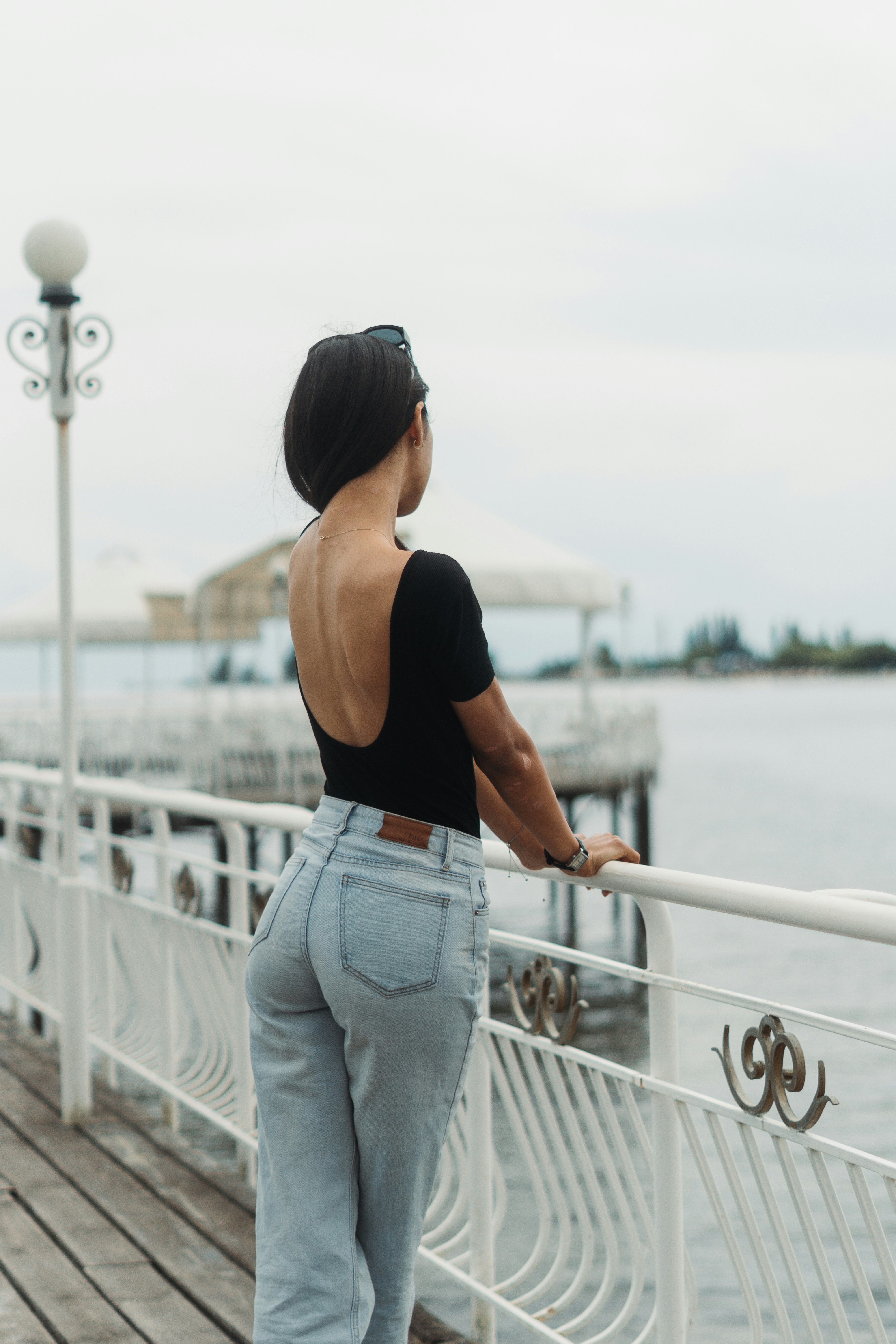 Woman in a backless top and light jeans gazing at the water from a wooden pier, with a soft focus on the serene surroundings.