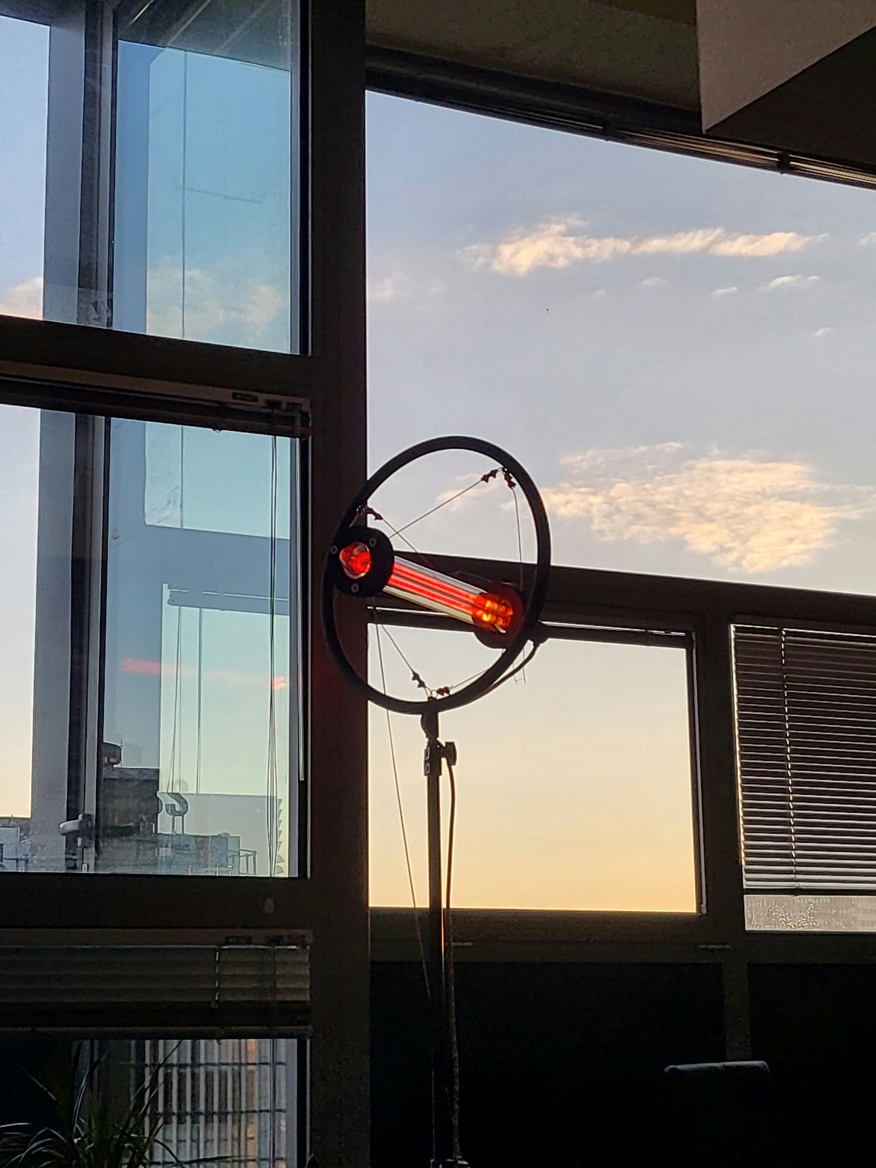Circular ring light with red accents dominates the foreground of a modern interior. A sunset gradient fills the windowed background.