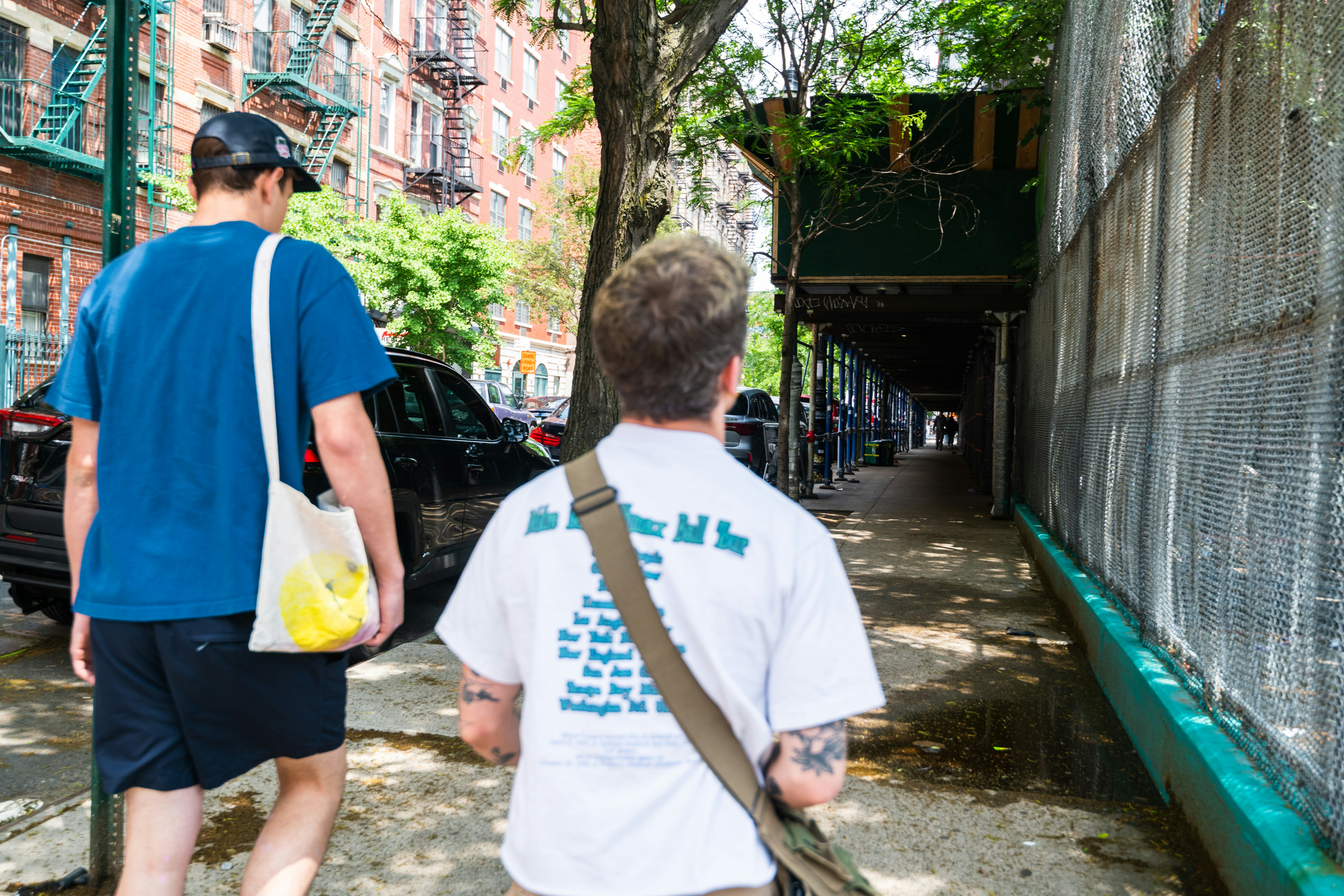 Two pedestrians along a sunlit alleyway with a chain-link fence on the right