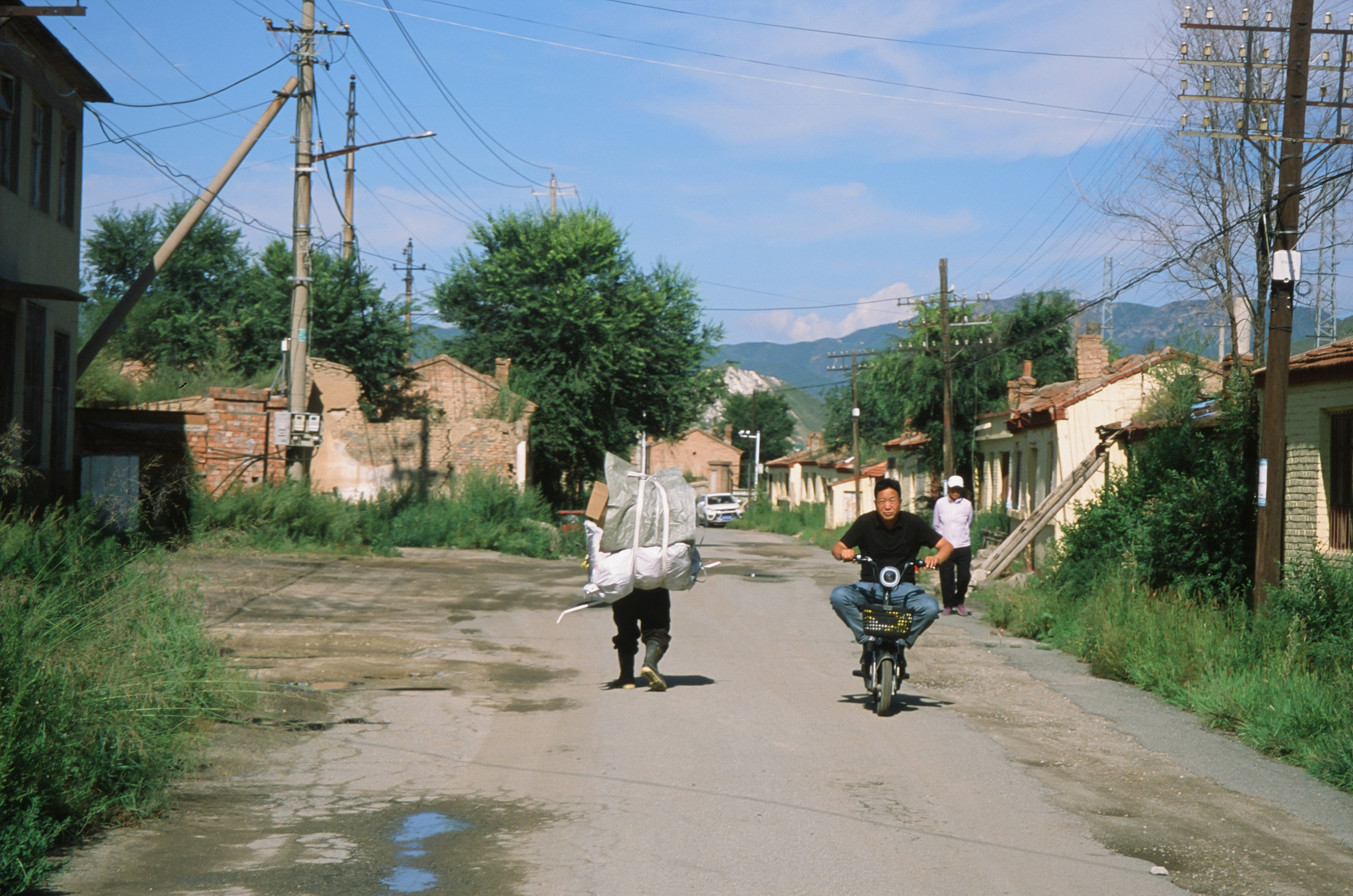 Sunlit rural street with a motorbike rider and a pedestrian crossing a weathered road, flanked by brick houses and green vegetation. Distant mountains frame the landscape beyond.