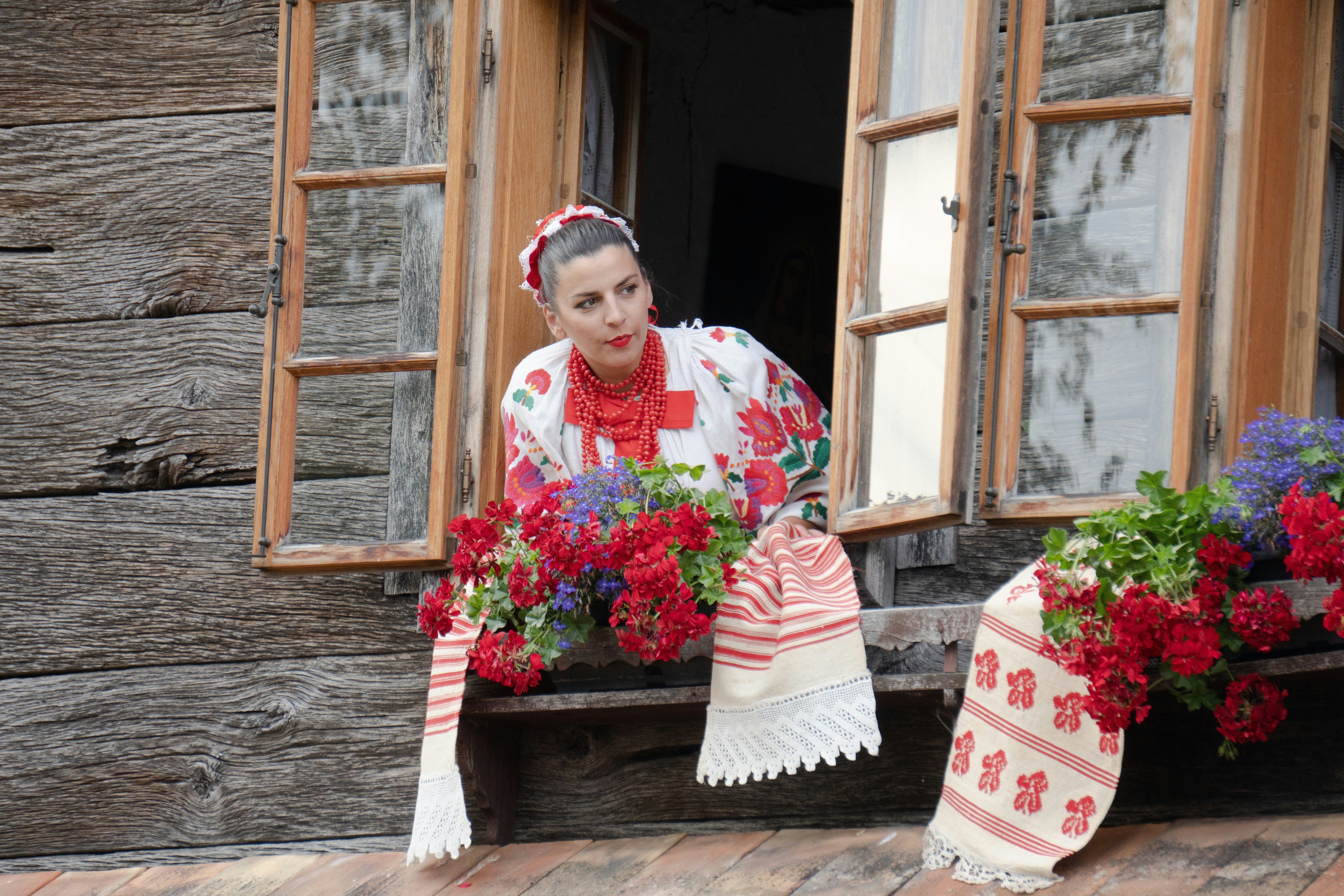 Woman in traditional attire peering out of a rustic window adorned with vibrant flowers and decorative textiles.