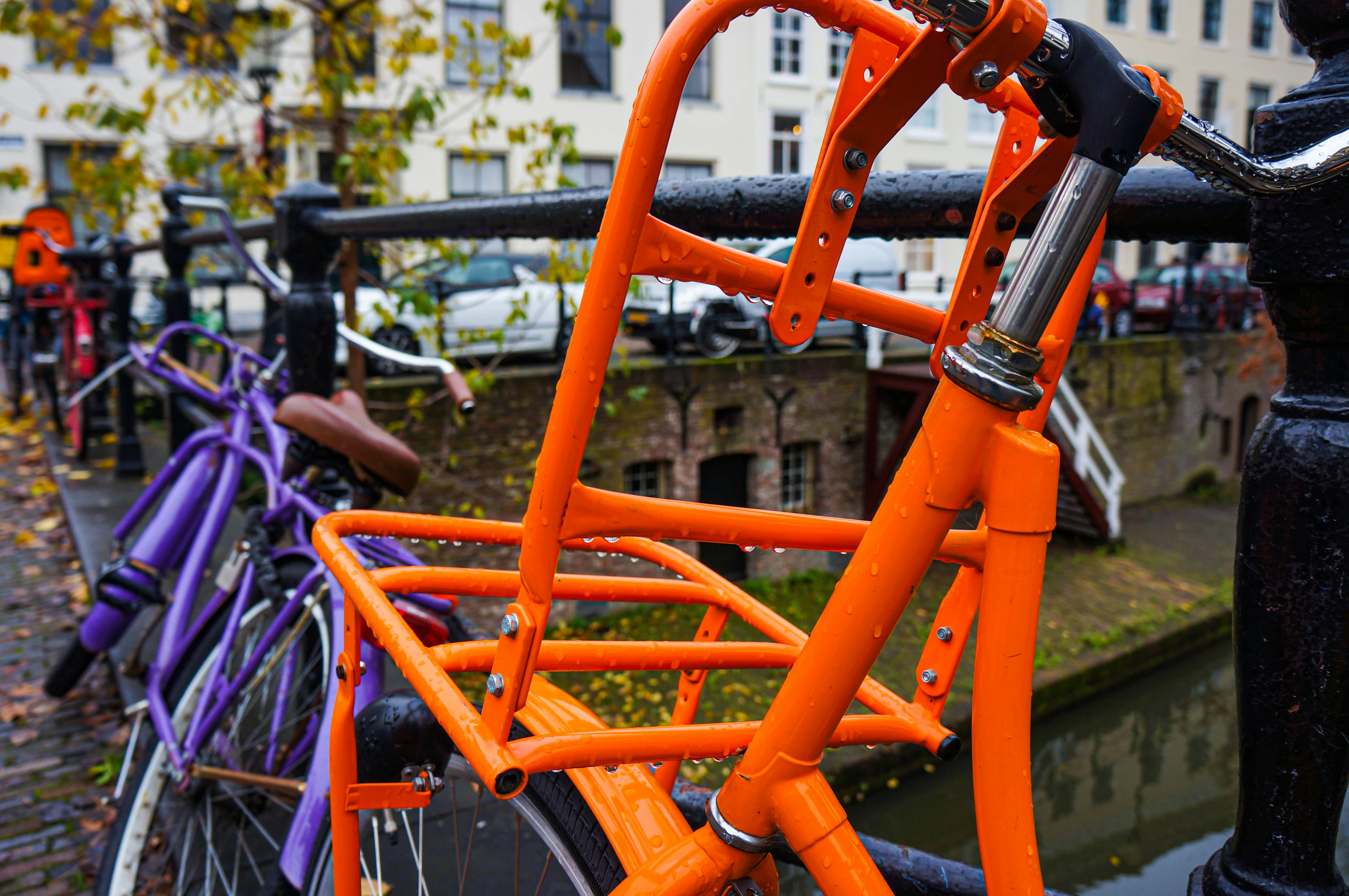 Orange bicycle frame in the foreground along a canal railing, with purple bikes fading into a brick backdrop.