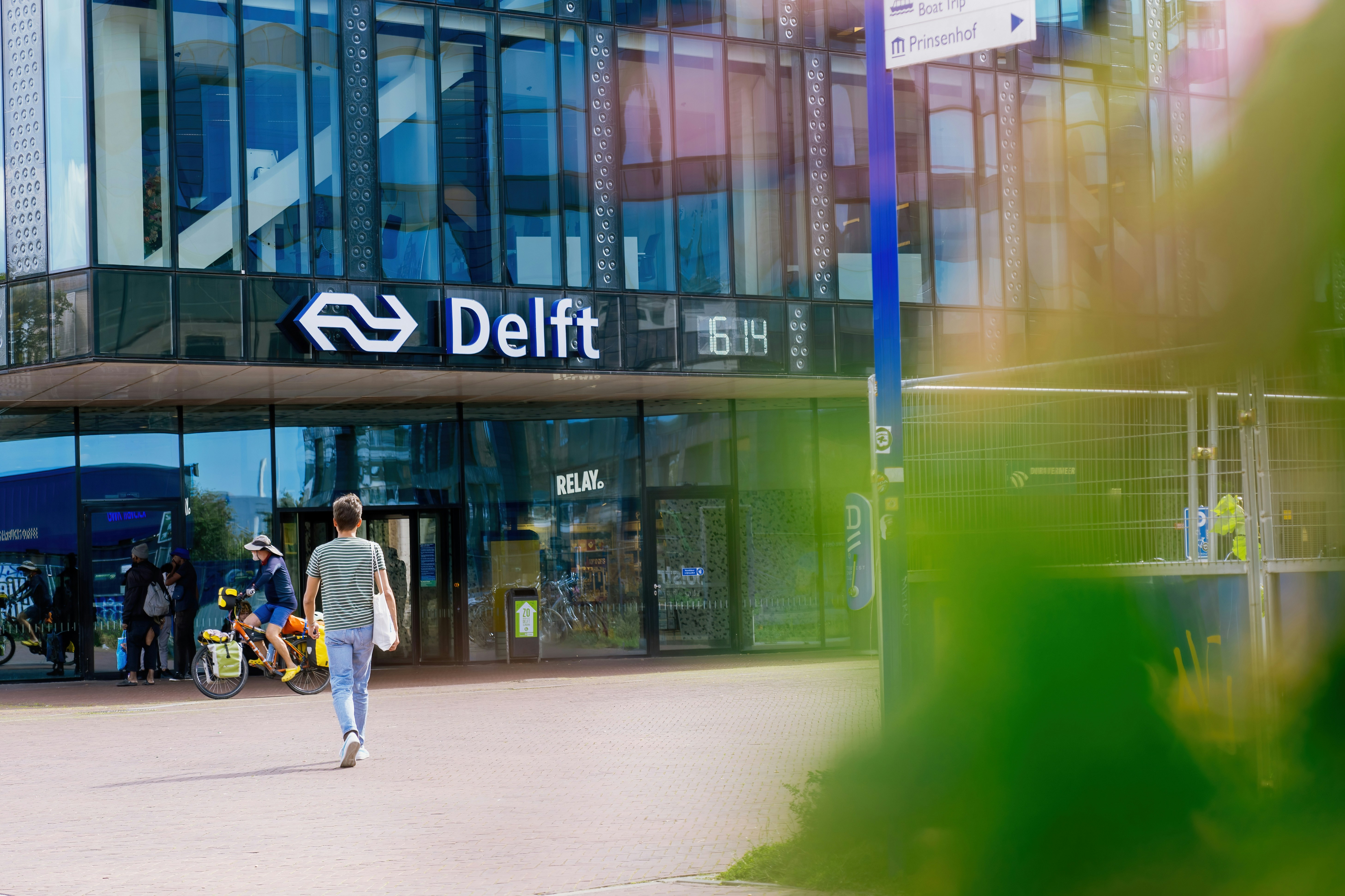 Person walking towards a modern building with 'Delft' sign, surrounded by green foliage and urban elements.